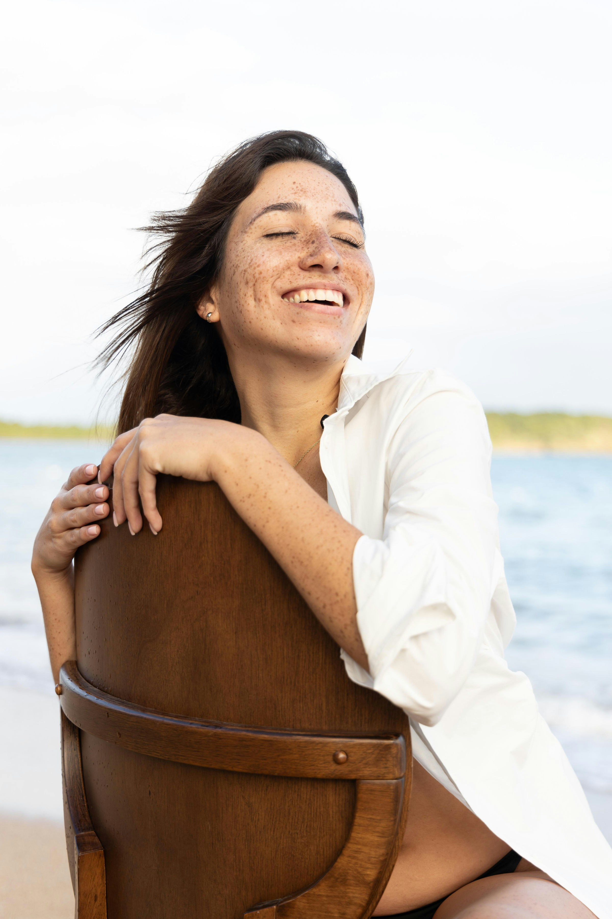 Bright portrait of a joyful woman leaning on a wooden chair by the seaside, hair fluttering and eyes closed in a carefree moment near the water.
