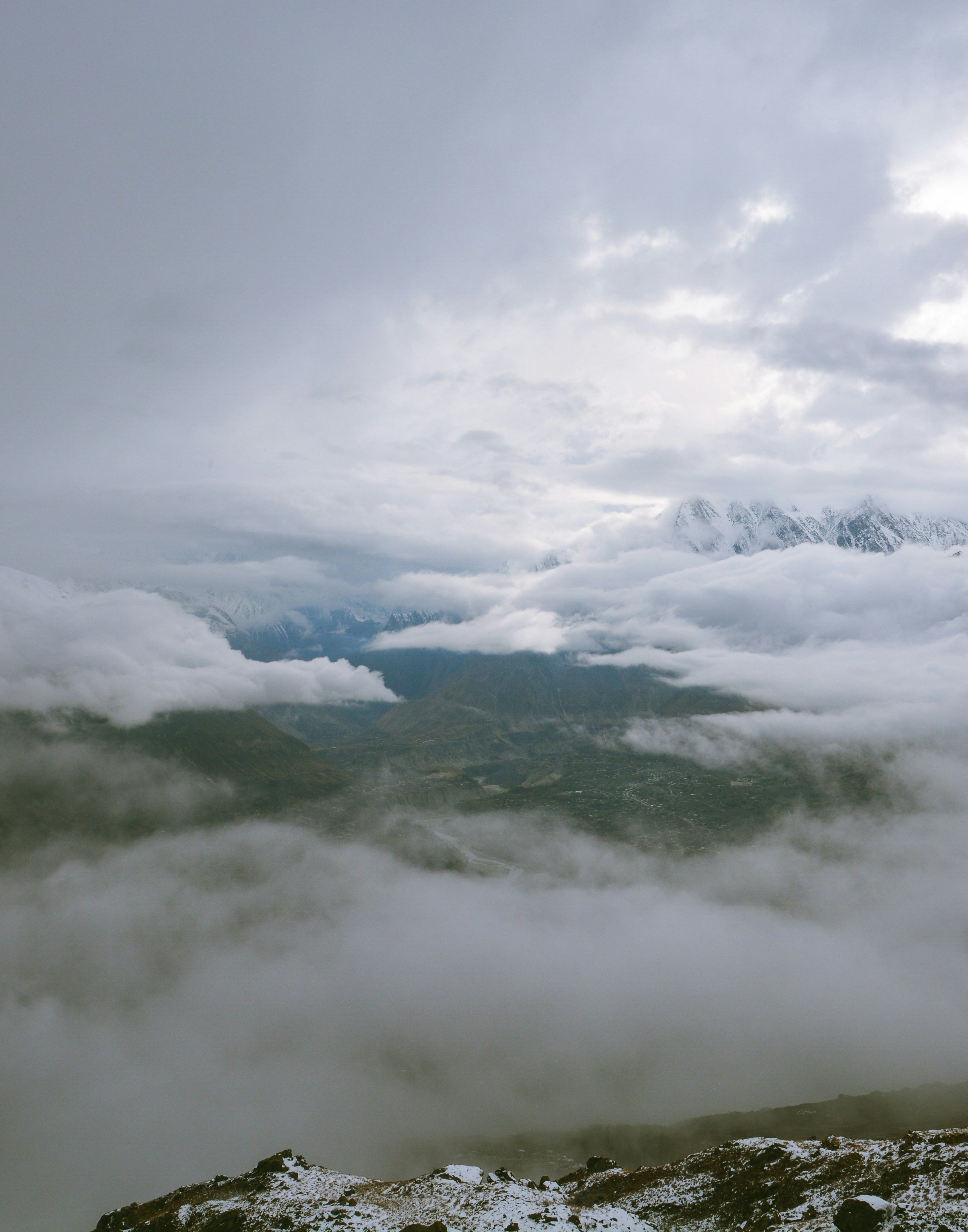 A high-elevation landscape shrouded in low-lying clouds and snow-capped ridges, viewed from above with a distant valley below.