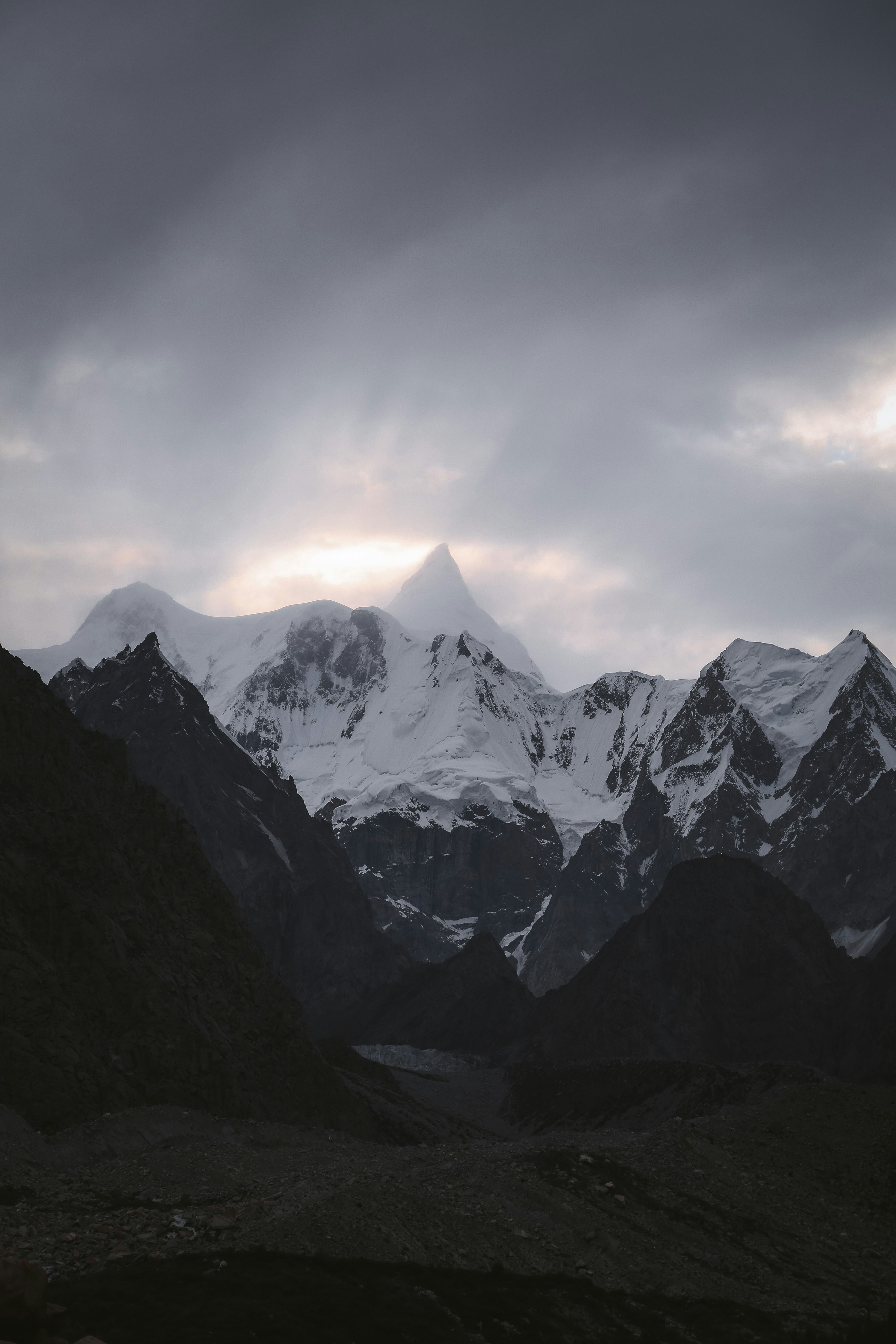 Snow-capped mountains loom beneath a moody, cloud-filled sky, with a dark valley in the foreground.
