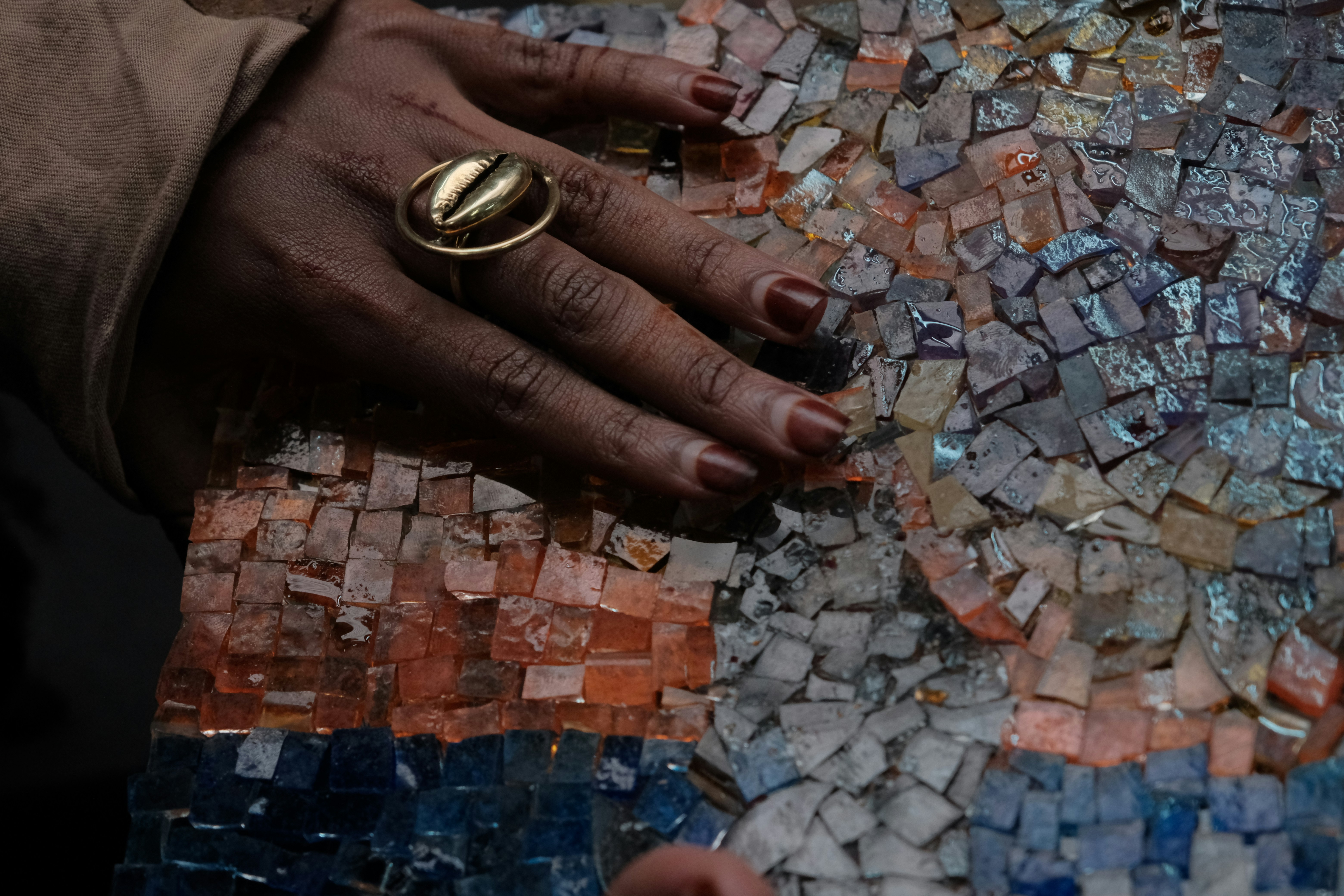 Close-up of a hand with a gold ring resting on a multicolor mosaic surface; the textures and hues of small square tiles create a rich, tactile backdrop.