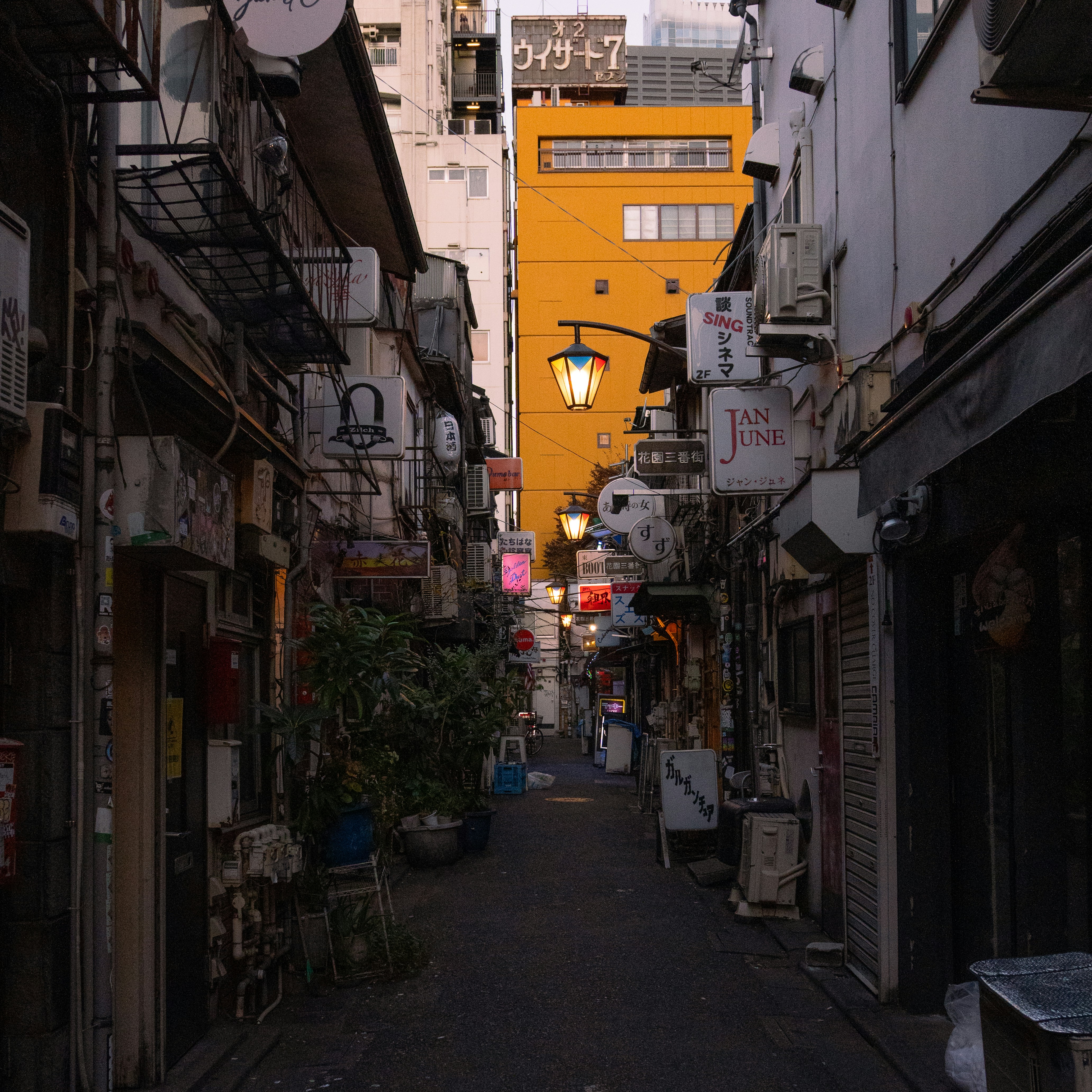 Narrow city alley with shops and signs lining both sides, leading to a brighter building at the end; ambient evening light and warm signage create a moody urban vignette.