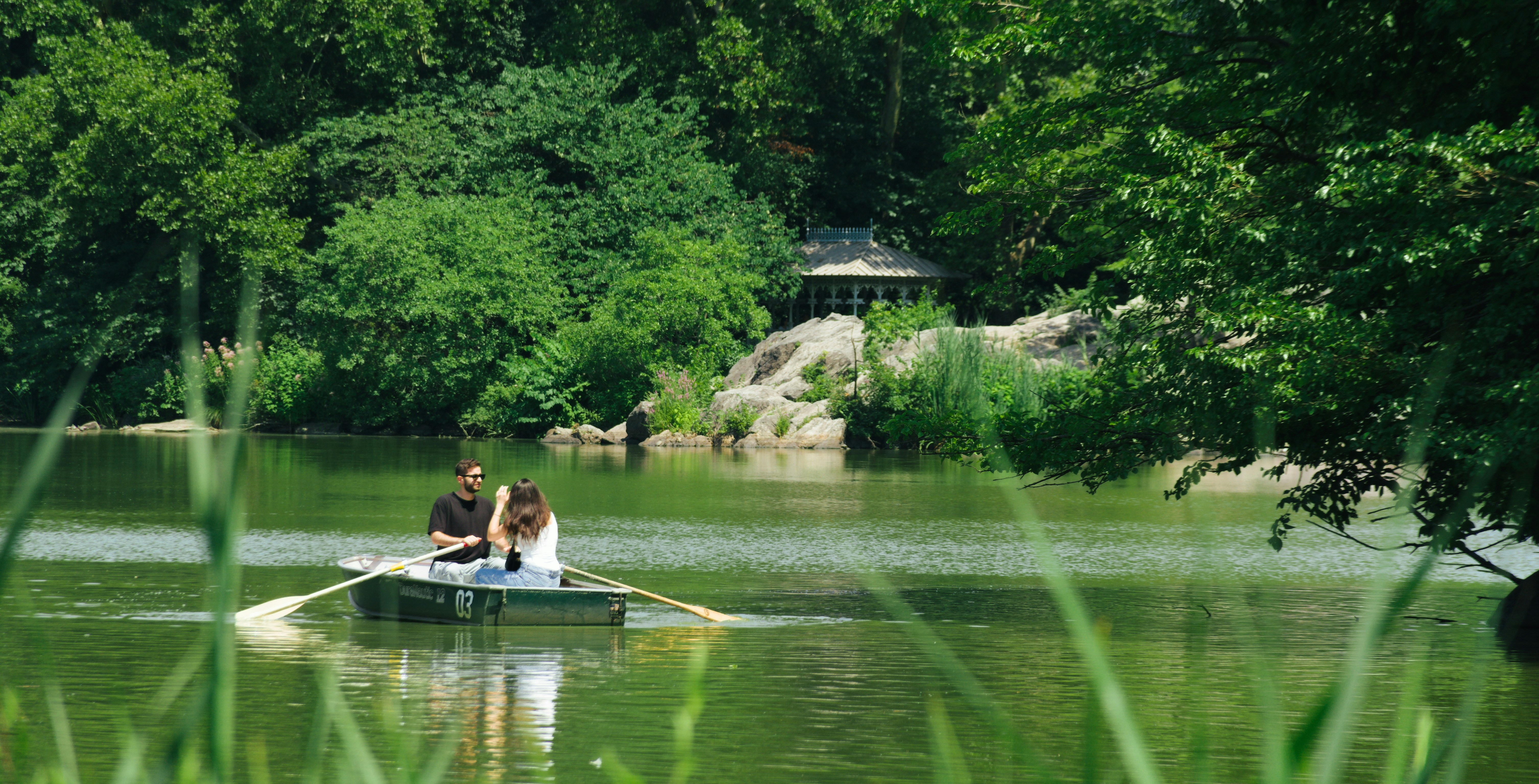 Two people rowing a small boat along a calm lakeside with a rocky, tree-lined shoreline and a small pavilion in the background.