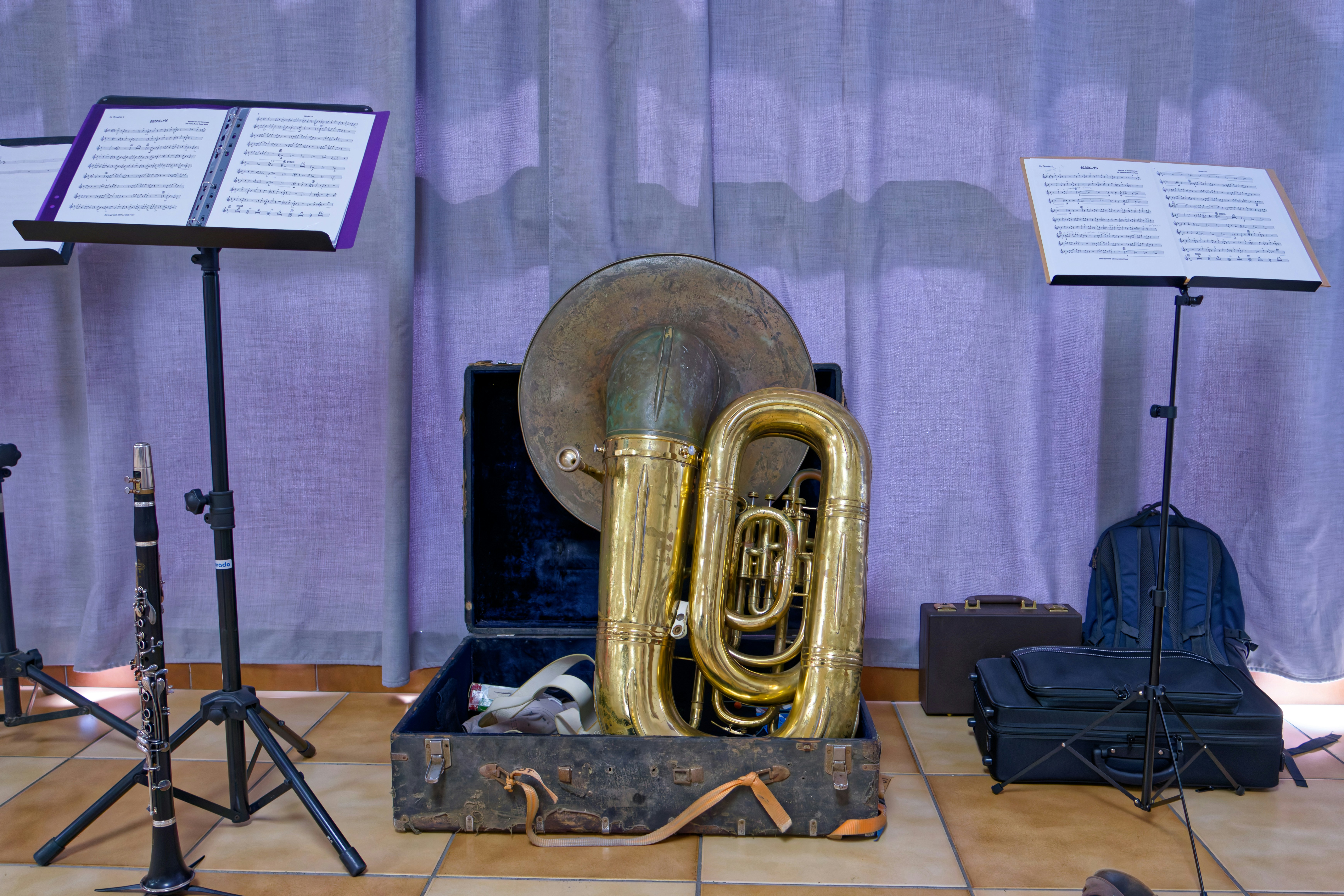 Brass instruments rest in an open case on a stage with music stands and purple curtains in the background.