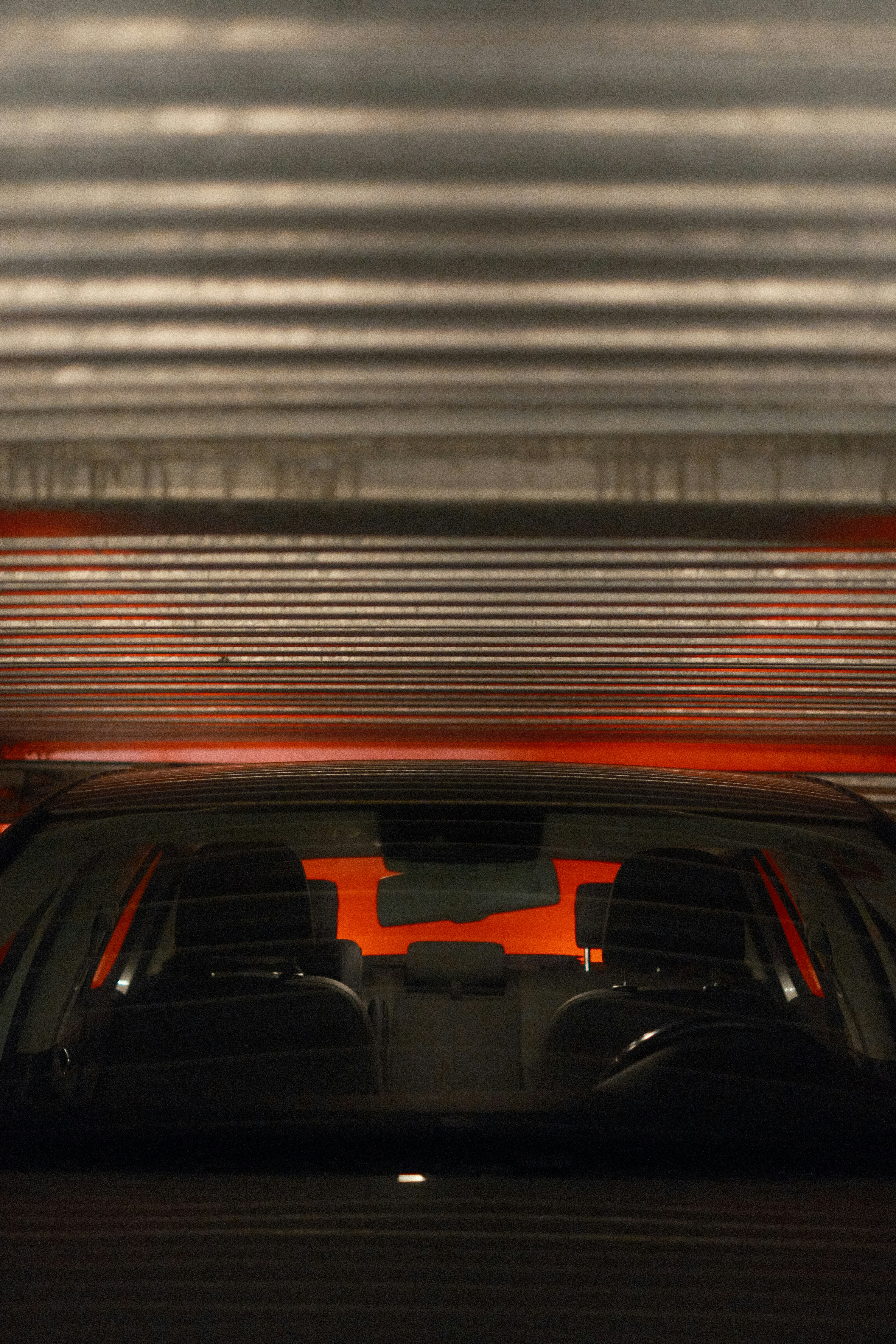 A red car is viewed from behind in a dim, striped ceiling-lit parking structure; interior seats and rear window glow softly through the glass.