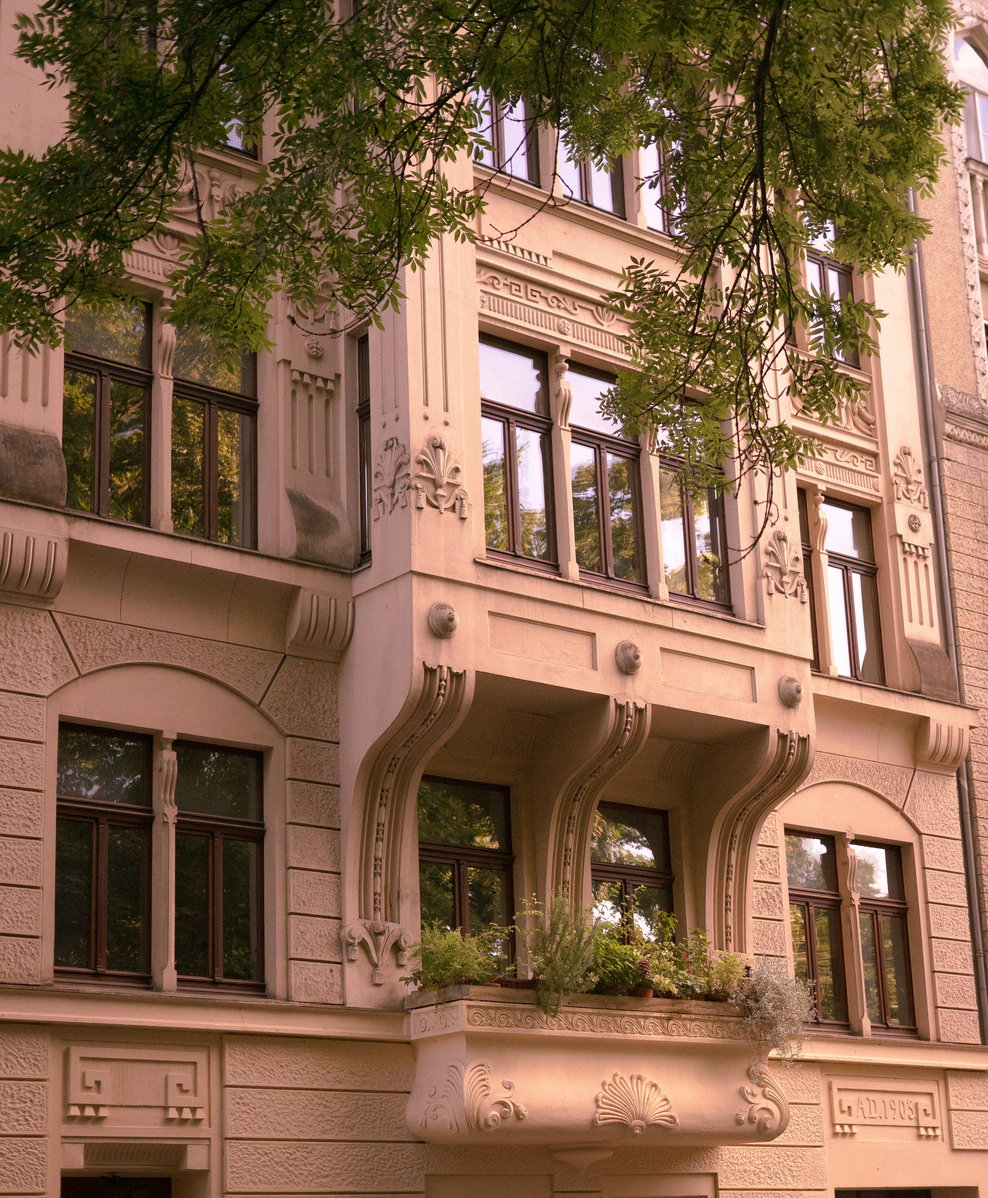An enchanting, dreamlike view of a historic Art Nouveau facade in Europe. Bathed in a warm, cinematic peach glow, the ornate stone carvings and elegant curves of the balcony are framed by soft green leaves, beautifully preserved from 1903 and evoking a sense of nostalgic poetry.