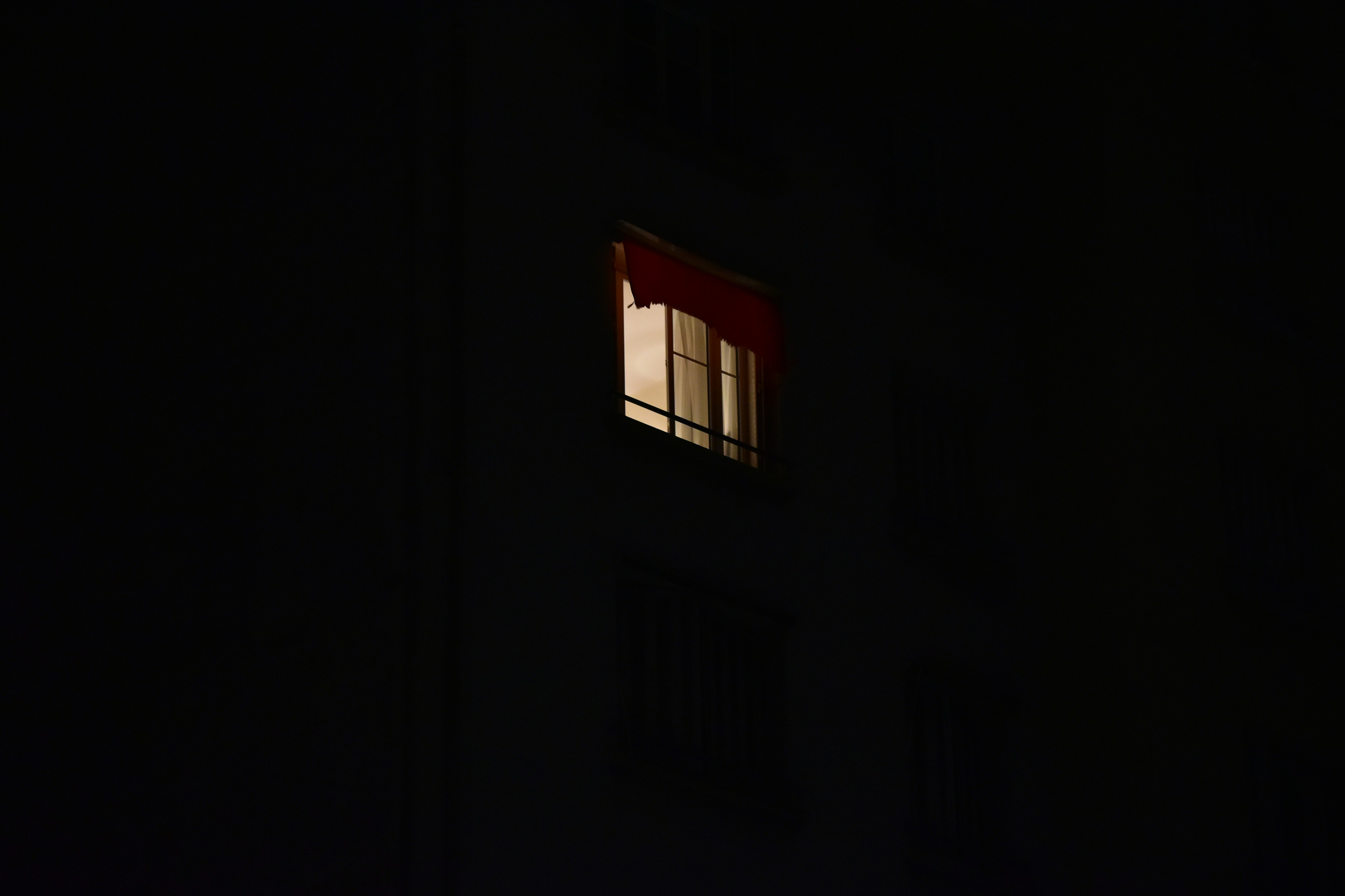 A single illuminated window glows softly from a dark, quiet building facade, hinting at activity within a night scene.