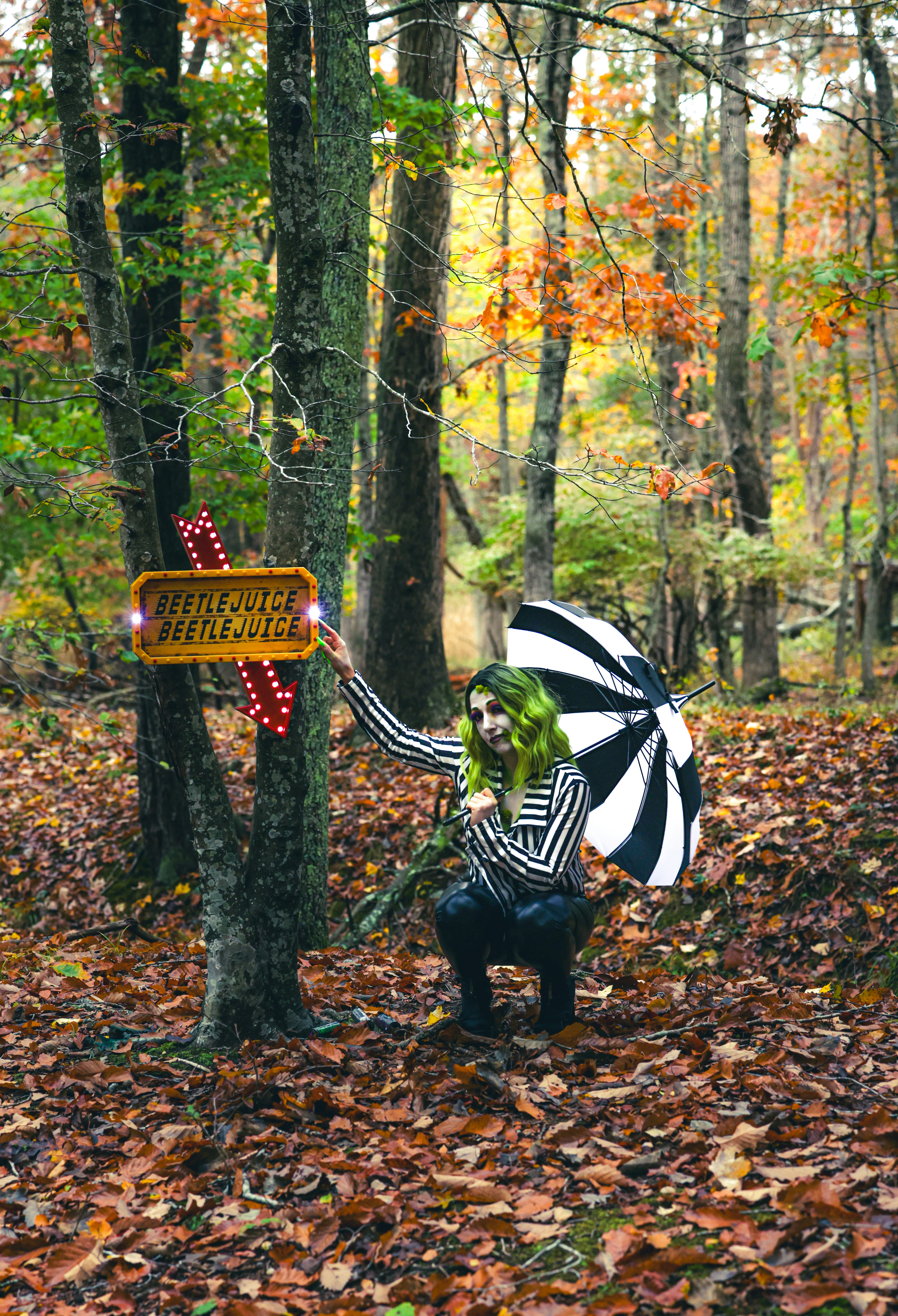 A model with lime-green hair crouches under a black-and-white umbrella among autumn leaves, pointing at a glowing sign in a forest setting.