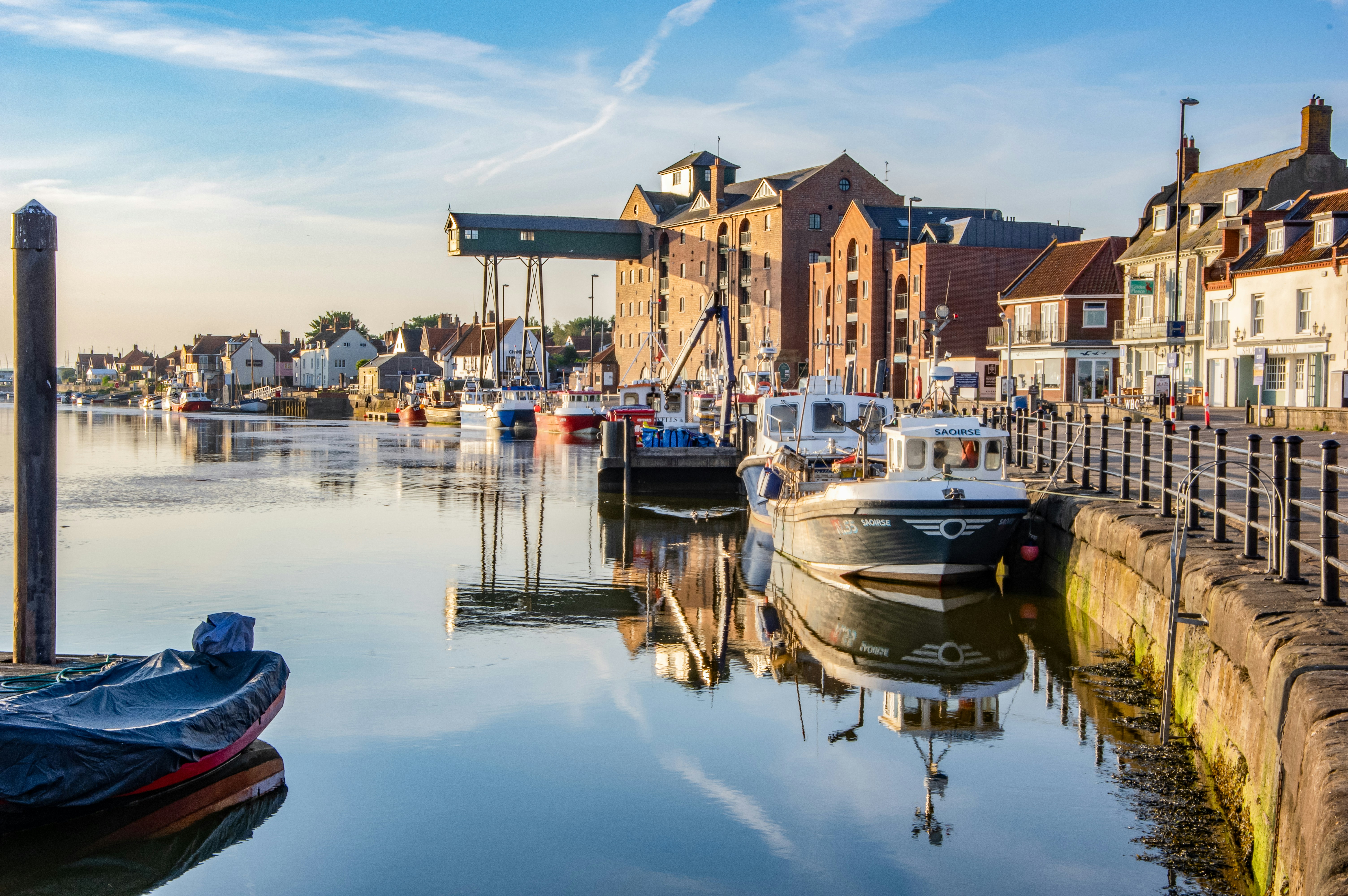 Calm harbor scene with boats moored along a stone quay and a row of brick buildings bathed in warm morning light.