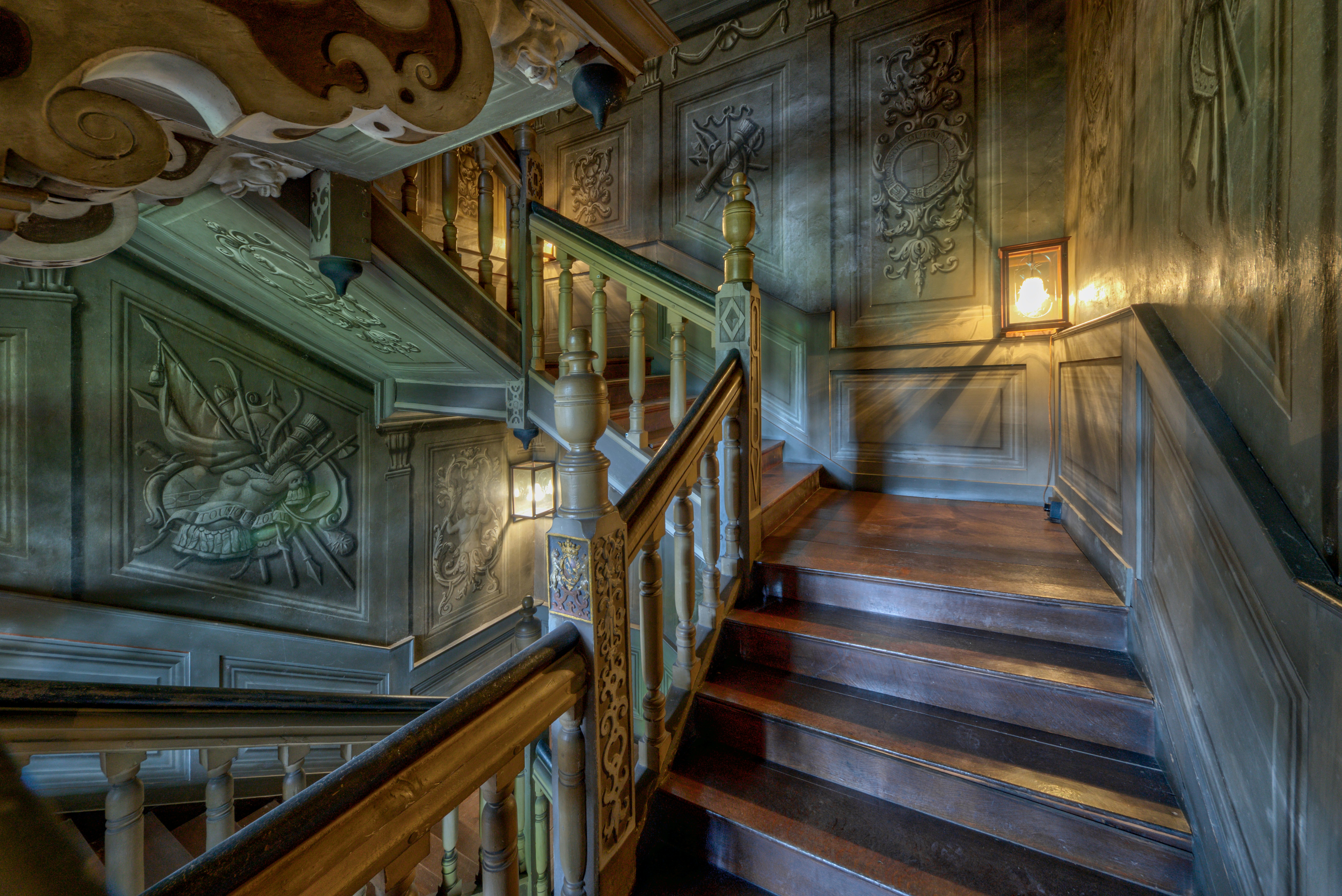 Carved wooden staircase winds upward in a richly detailed, orange-wold interior with ornate green walls and glowing wall sconces.