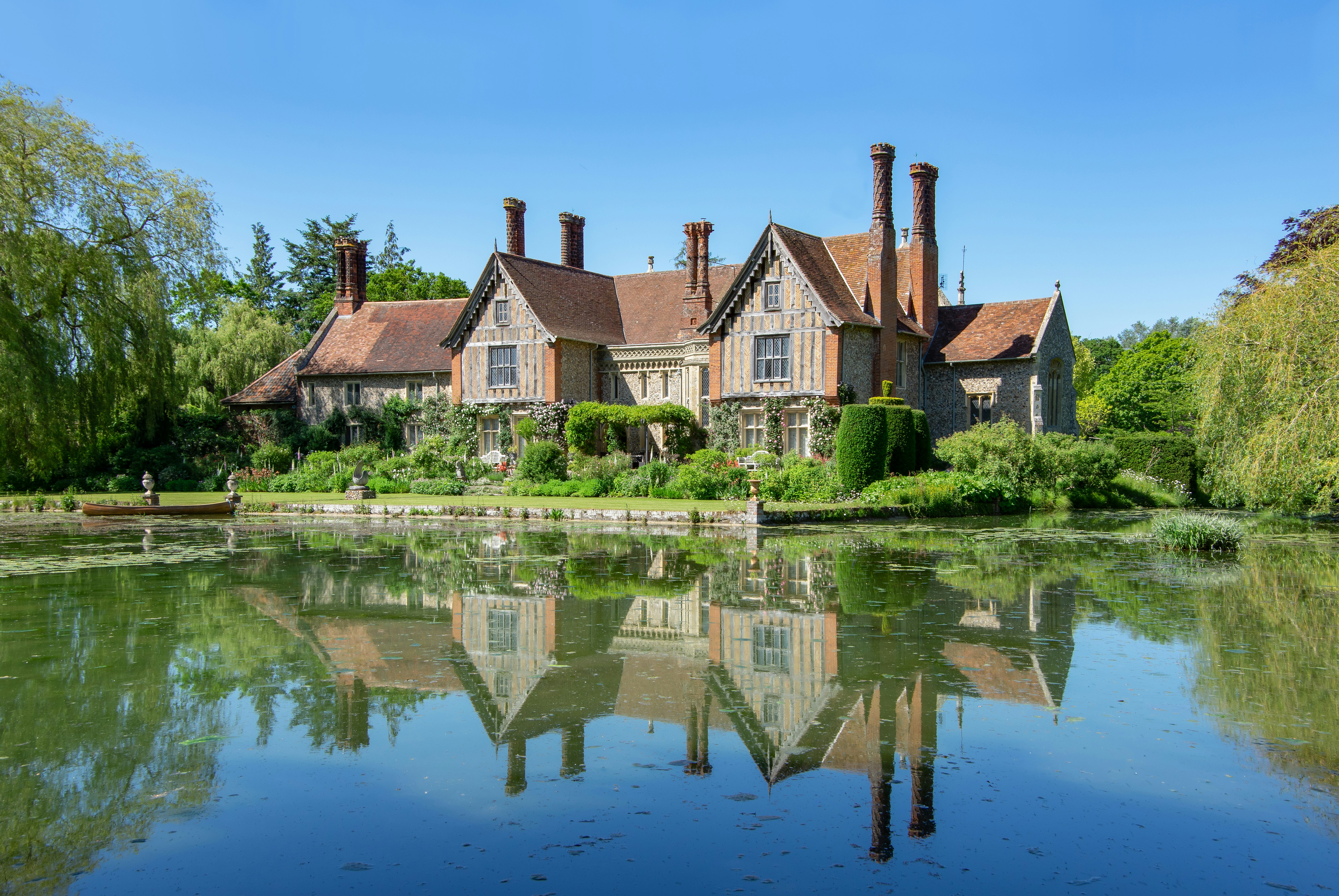 A stately manor sits beside a calm lake, its reflection perfectly mirrored in the water, with manicured gardens and a bright blue sky in the background.