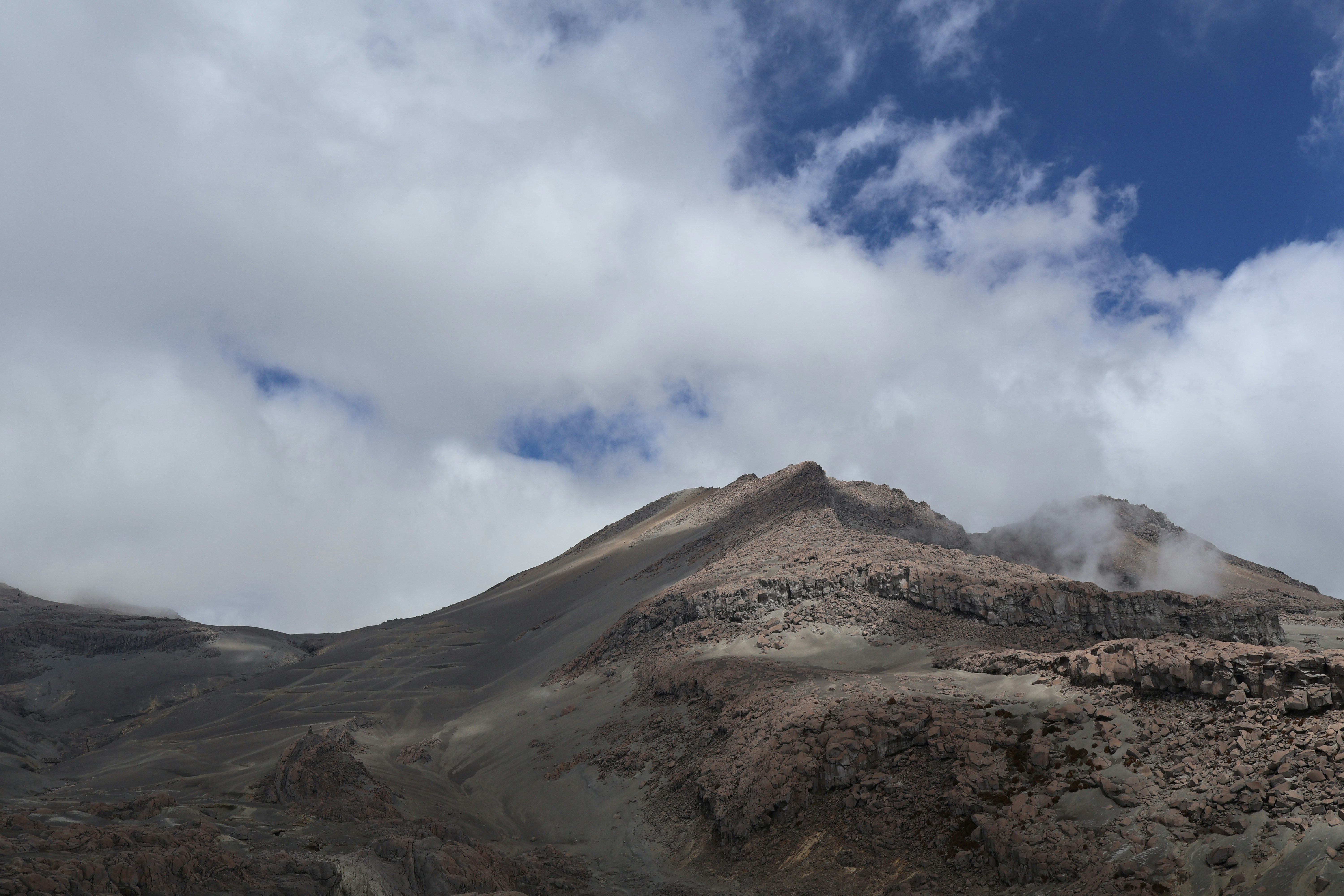Charred volcanic landscape with a distant peak rising above rugged lava fields under a partly cloudy sky. A stark, desolate panorama highlighting the terrain's textures and geological layers.