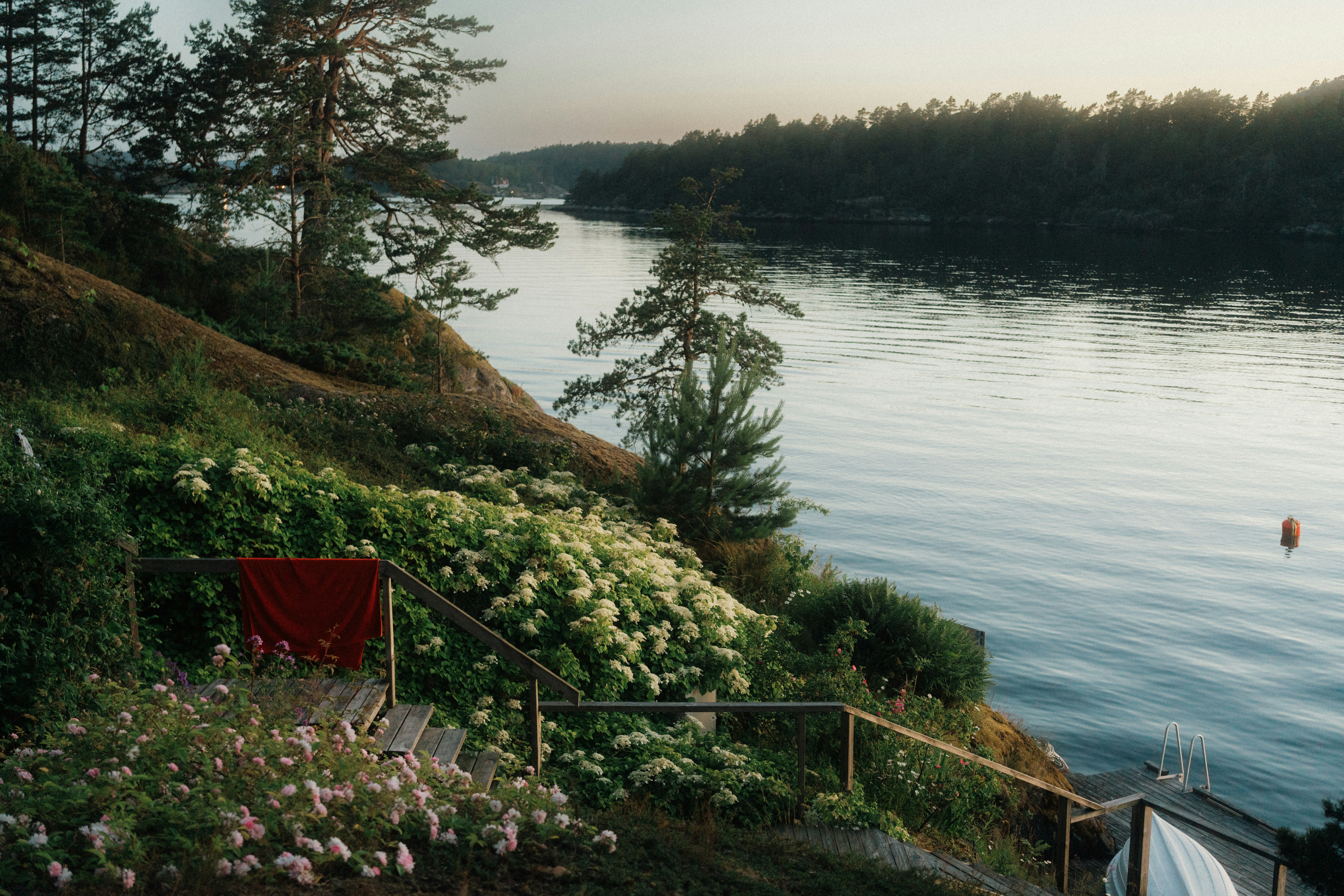 Colorful garden steps and flowering shrubs descend toward a calm lake framed by trees; a red cloth on a railing hints at a small, quiet cove by the water.