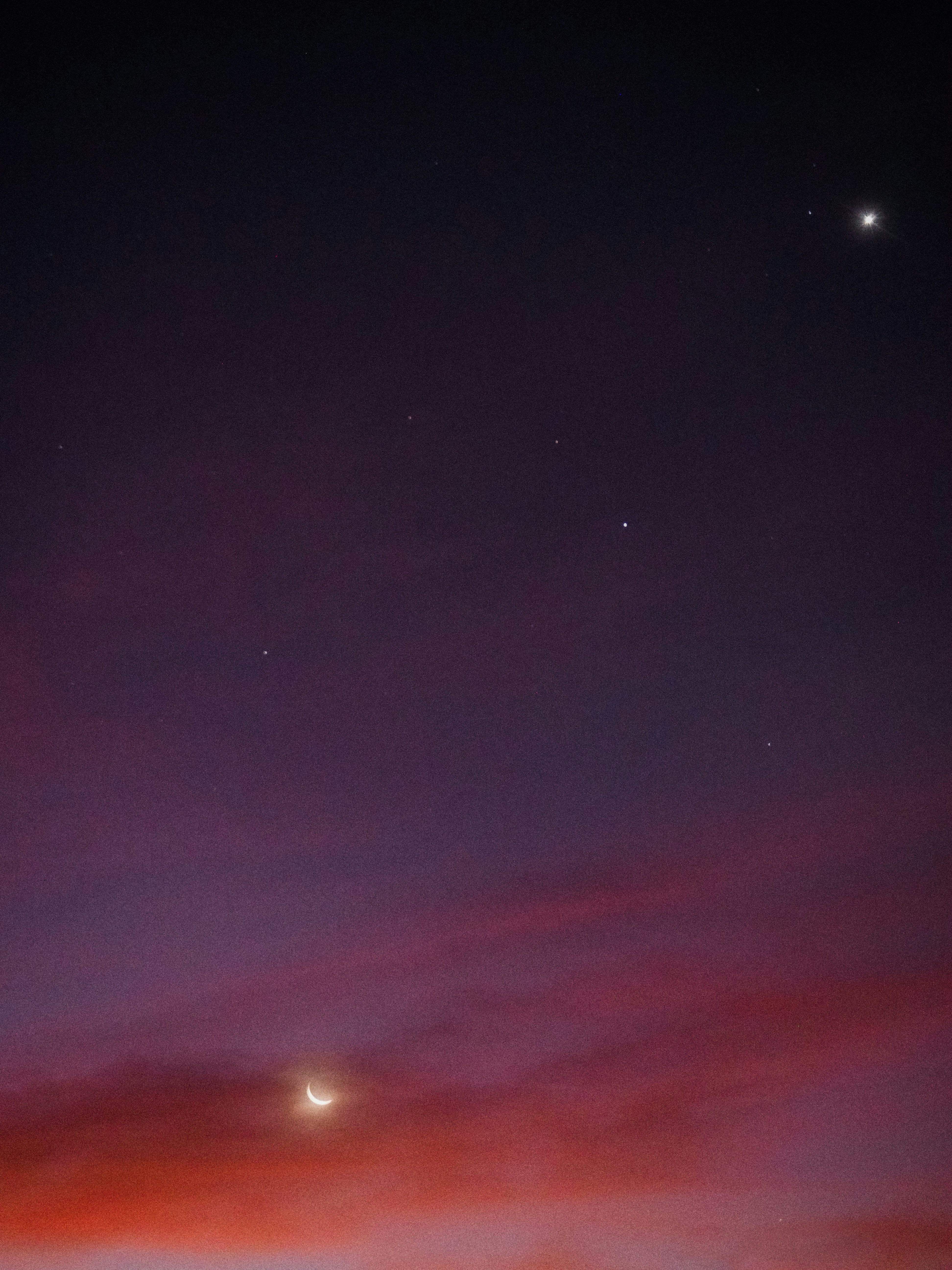 A slender crescent moon rises above a layered red-orange horizon, dotted with a few stars as the sky transitions from deep purple to warm crimson.