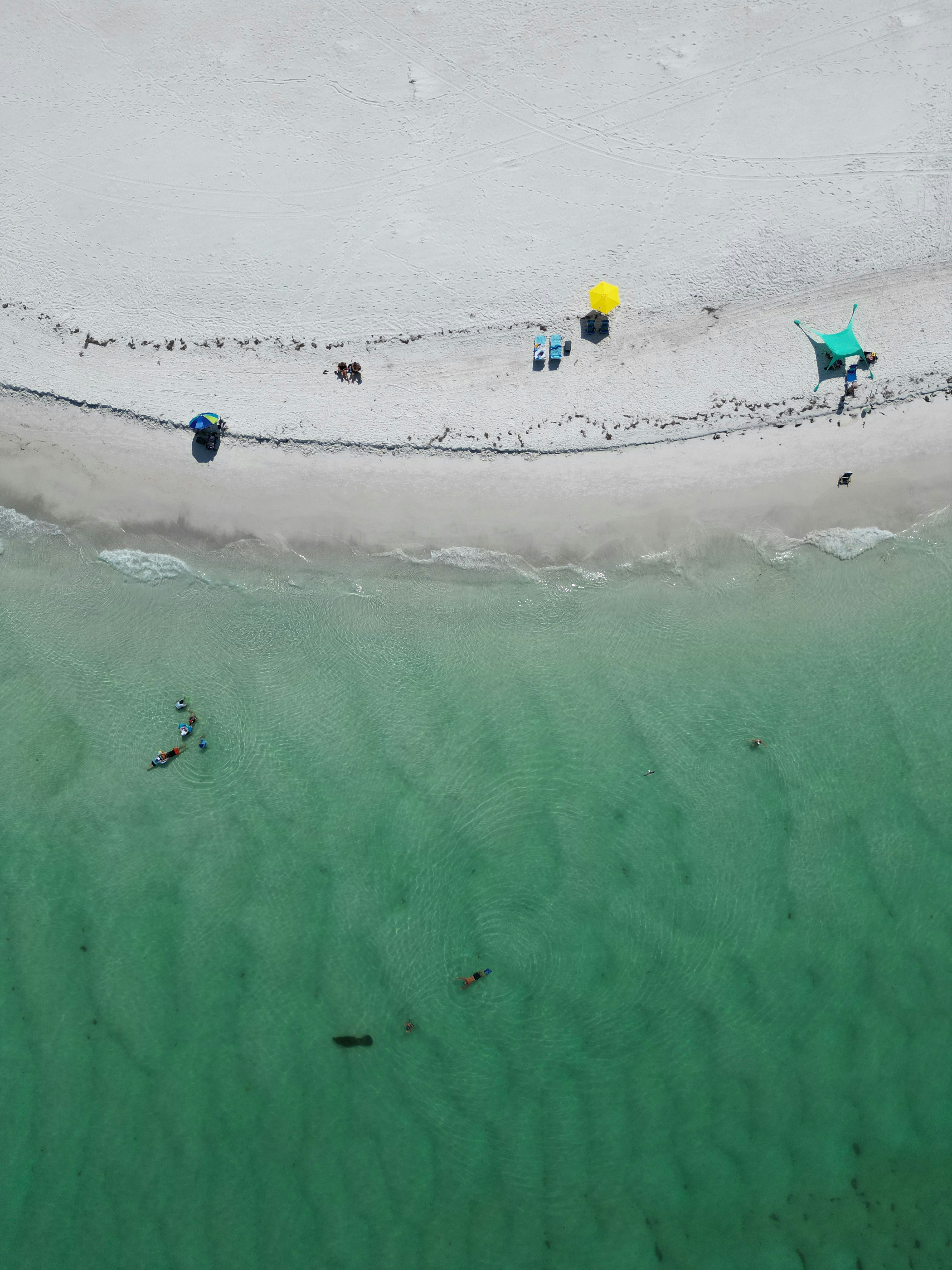 Bright beach scene captured from above showing shoreline, turquoise sea, and a few beachgoers with colorful parasols.