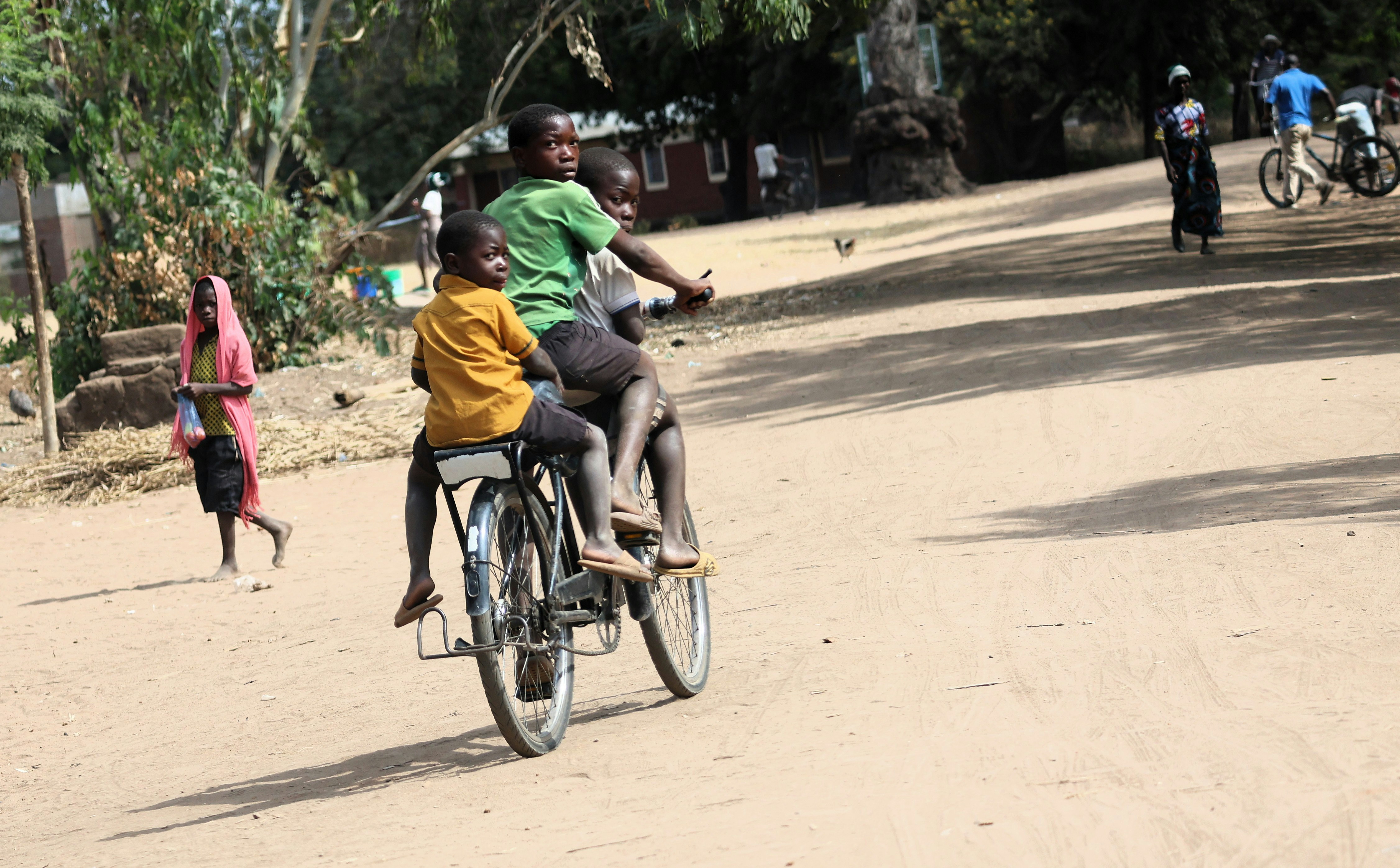Three kids ride a bicycle across a dusty street, with a girl in a pink veil watching, and other figures in the background; a simple village scene with natural light.