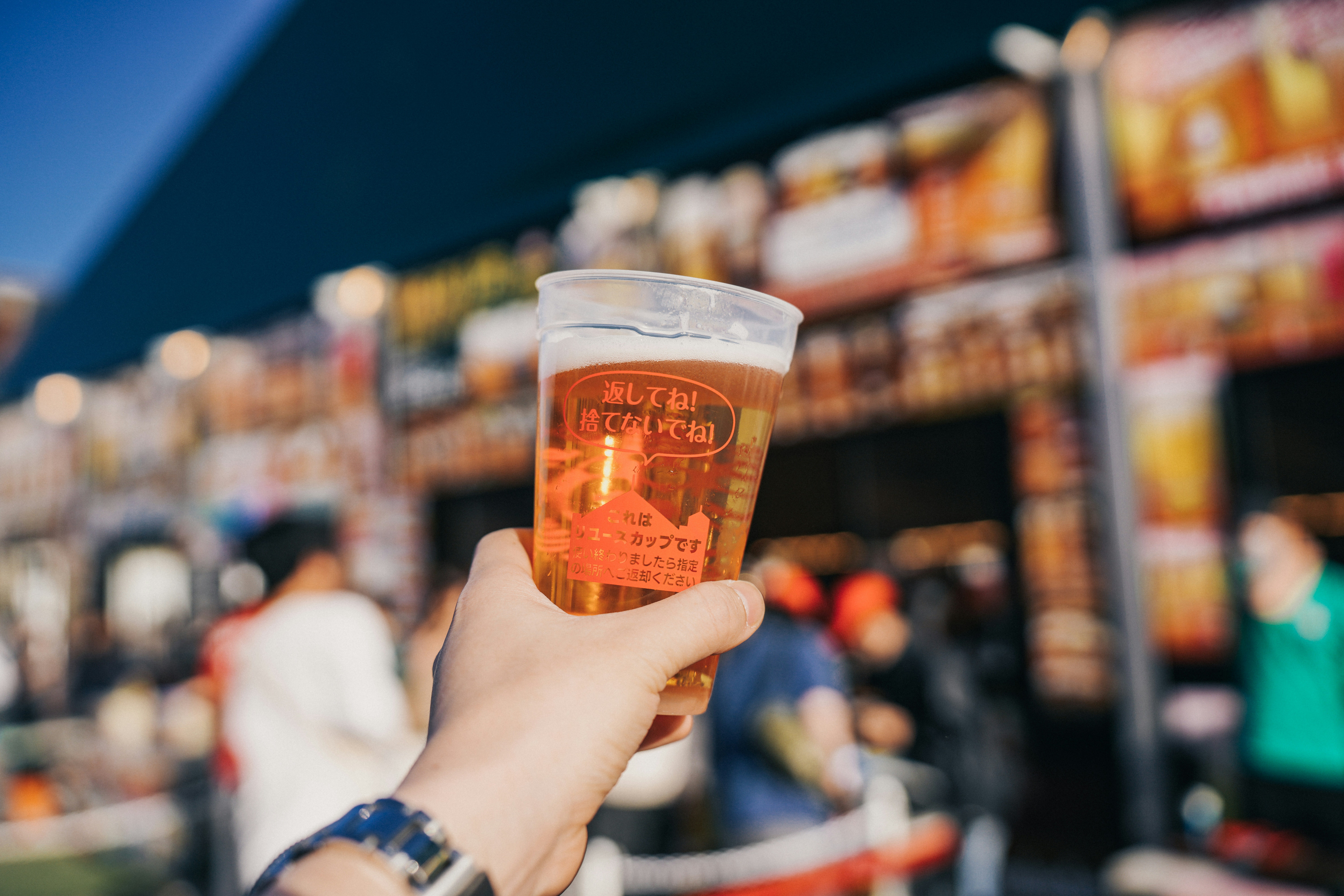 A hand holding a cup of beer foregrounds a lively, vendor-filled scene with blurred festival lights in the background.