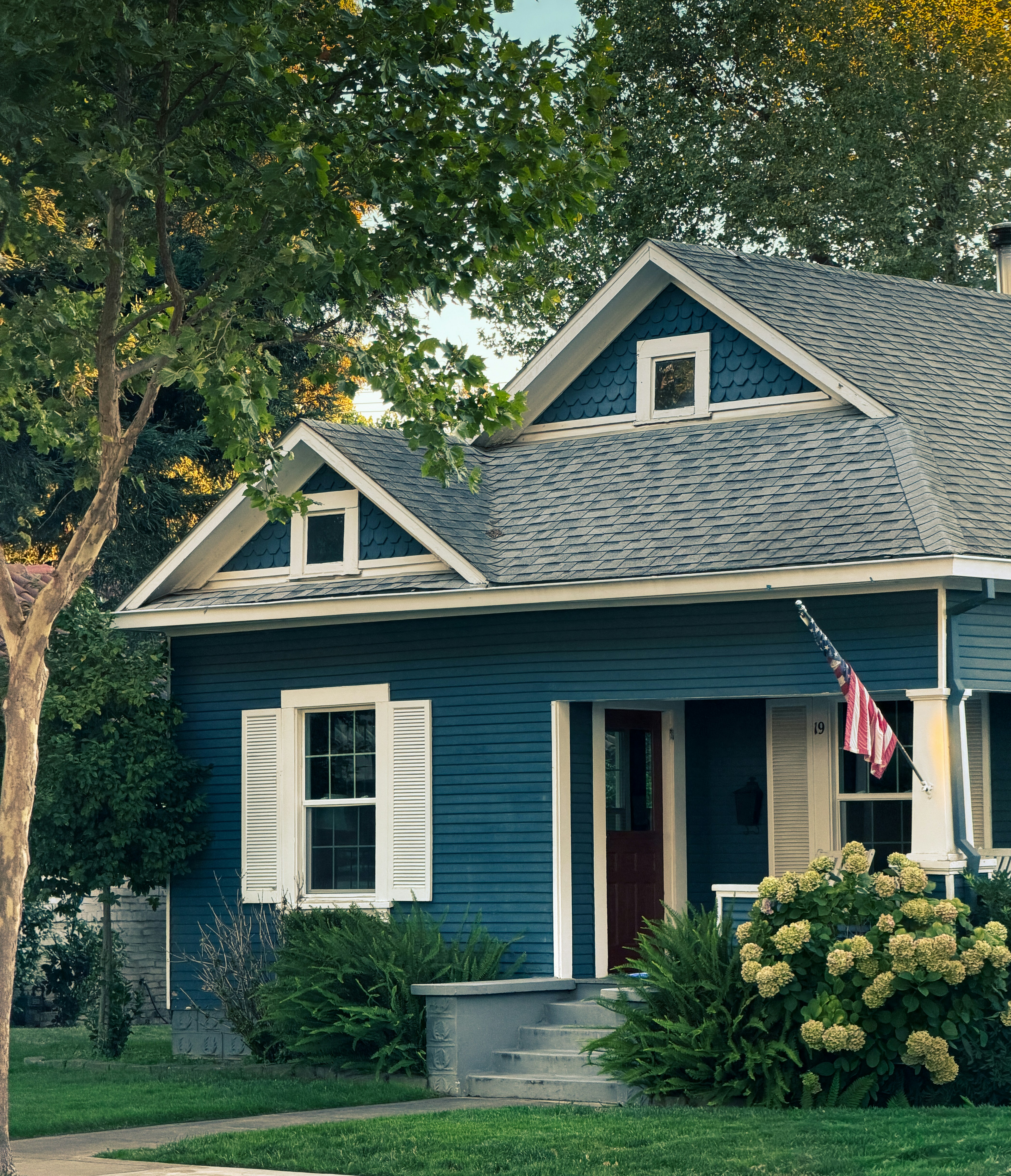 Blue wooden house with white trim, porch, and American flag; lush landscaping and a large tree frame the scene in warm daylight.