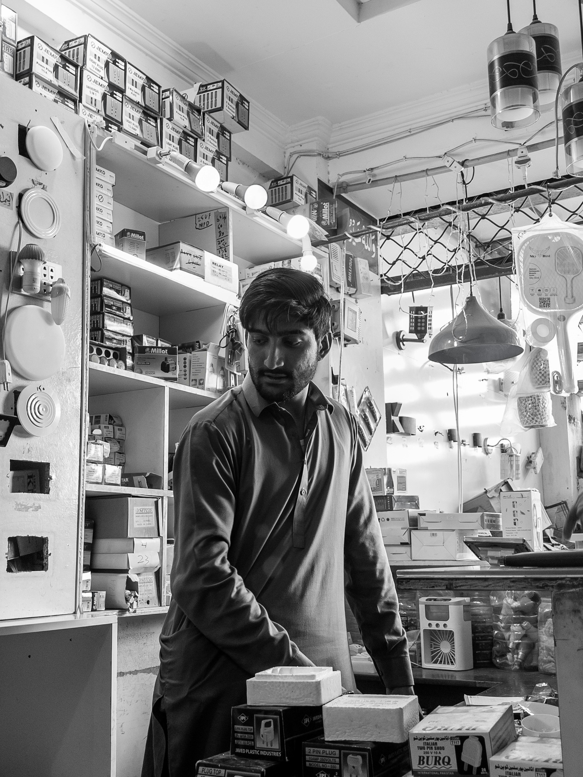 A grayscale scene inside a cluttered shop shows a man in a kurta arranging boxes, shelves filled with goods and equipment in the background.