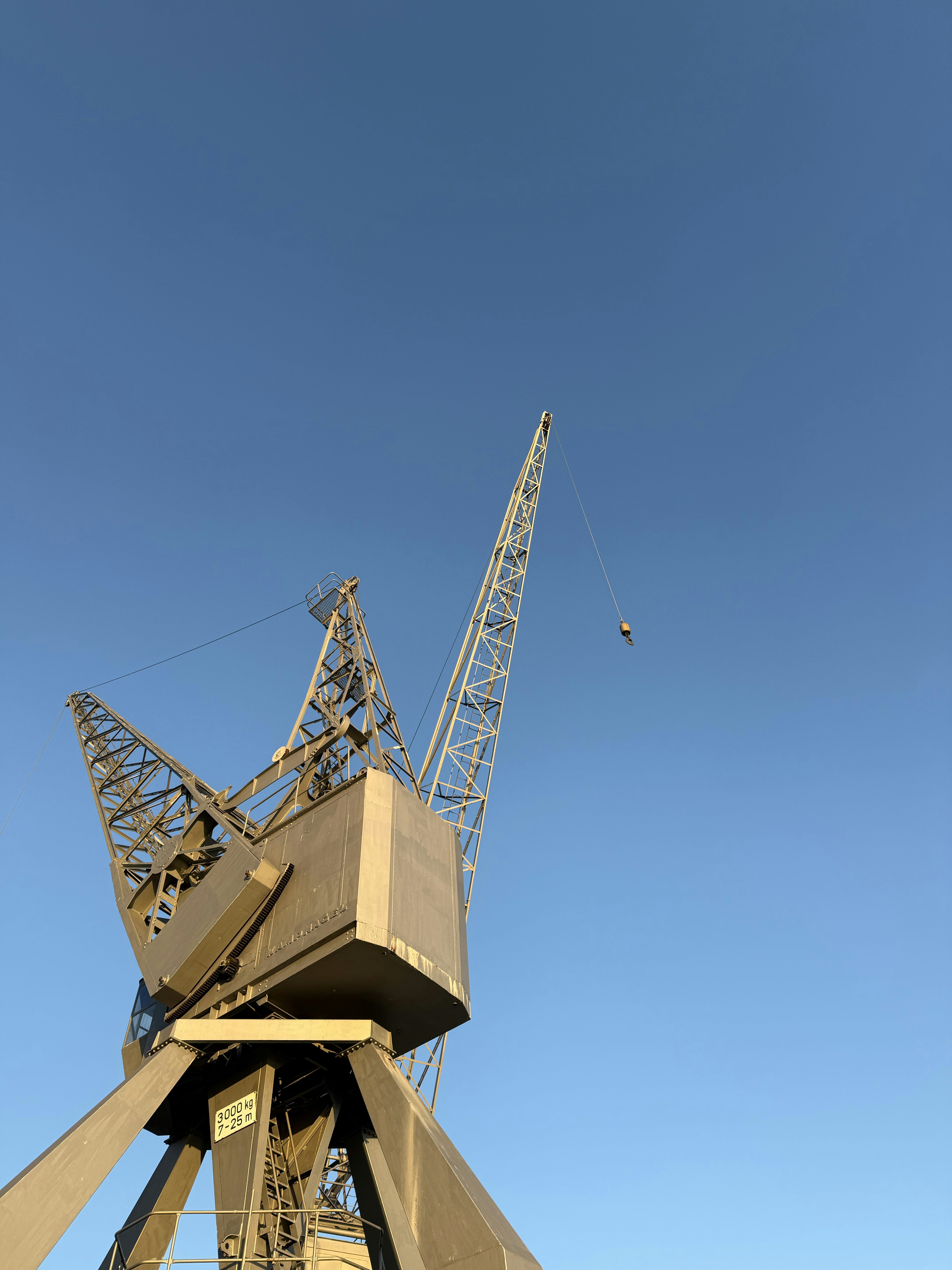 Industrial crane silhouette against a clear blue sky, with structural details and a hanging cable in the upper right; a sturdy metal framework dominates the lower left, conveying scale and elevation.