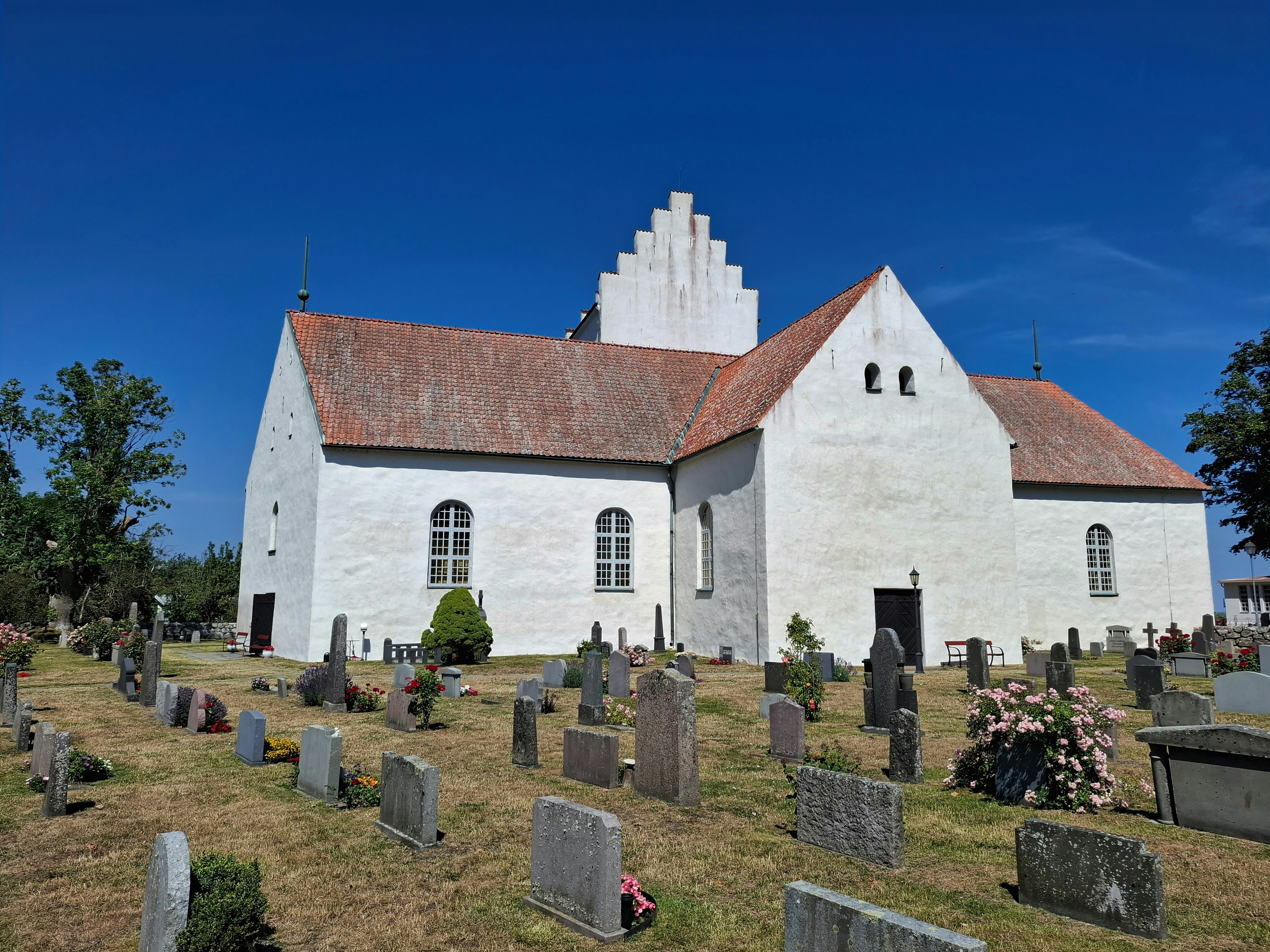 White church with red roof sits among a graveyard; rows of headstones and flowering shrubs extend toward a bright blue sky, conveying a serene rural setting.