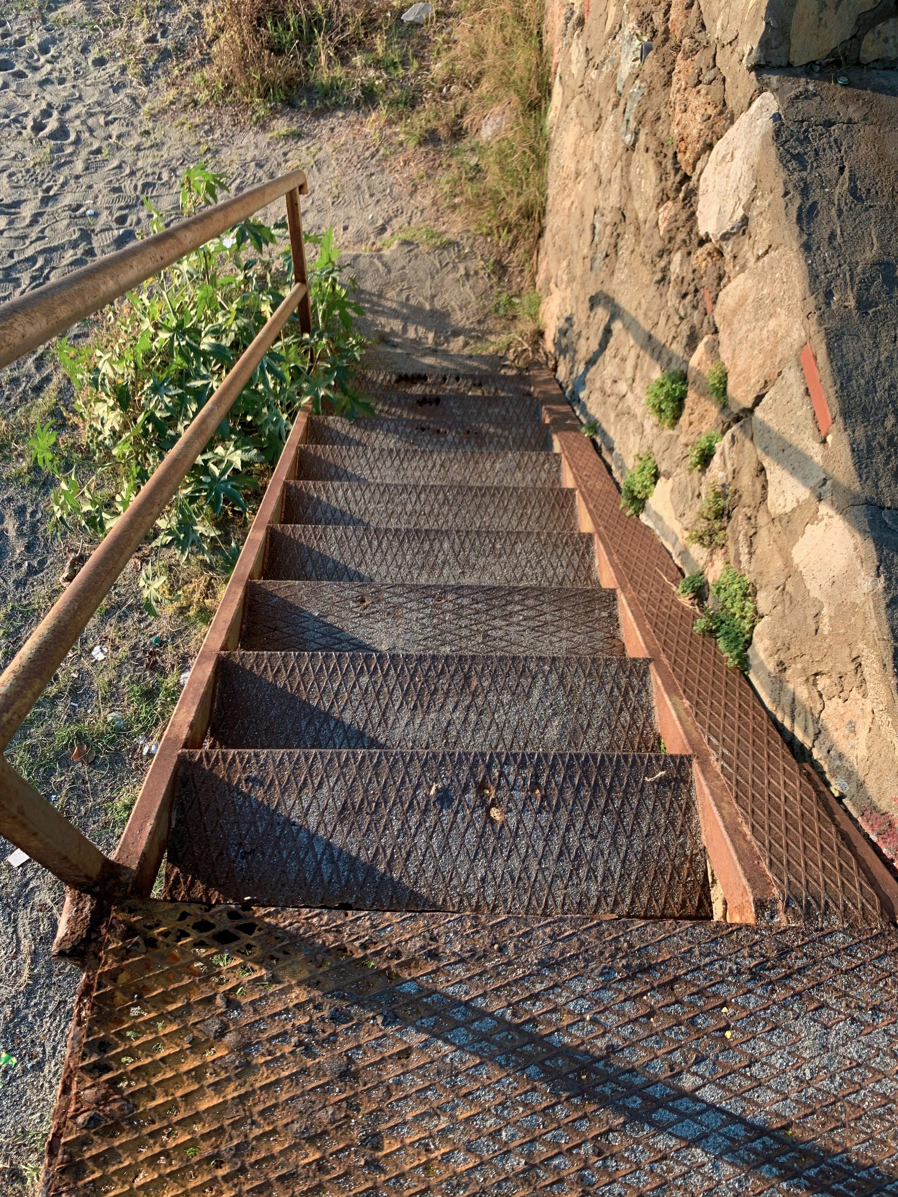 Rusty metal stairs descend along a stone wall toward a rocky ground, with a handrail casting a shadow on the grated steps.