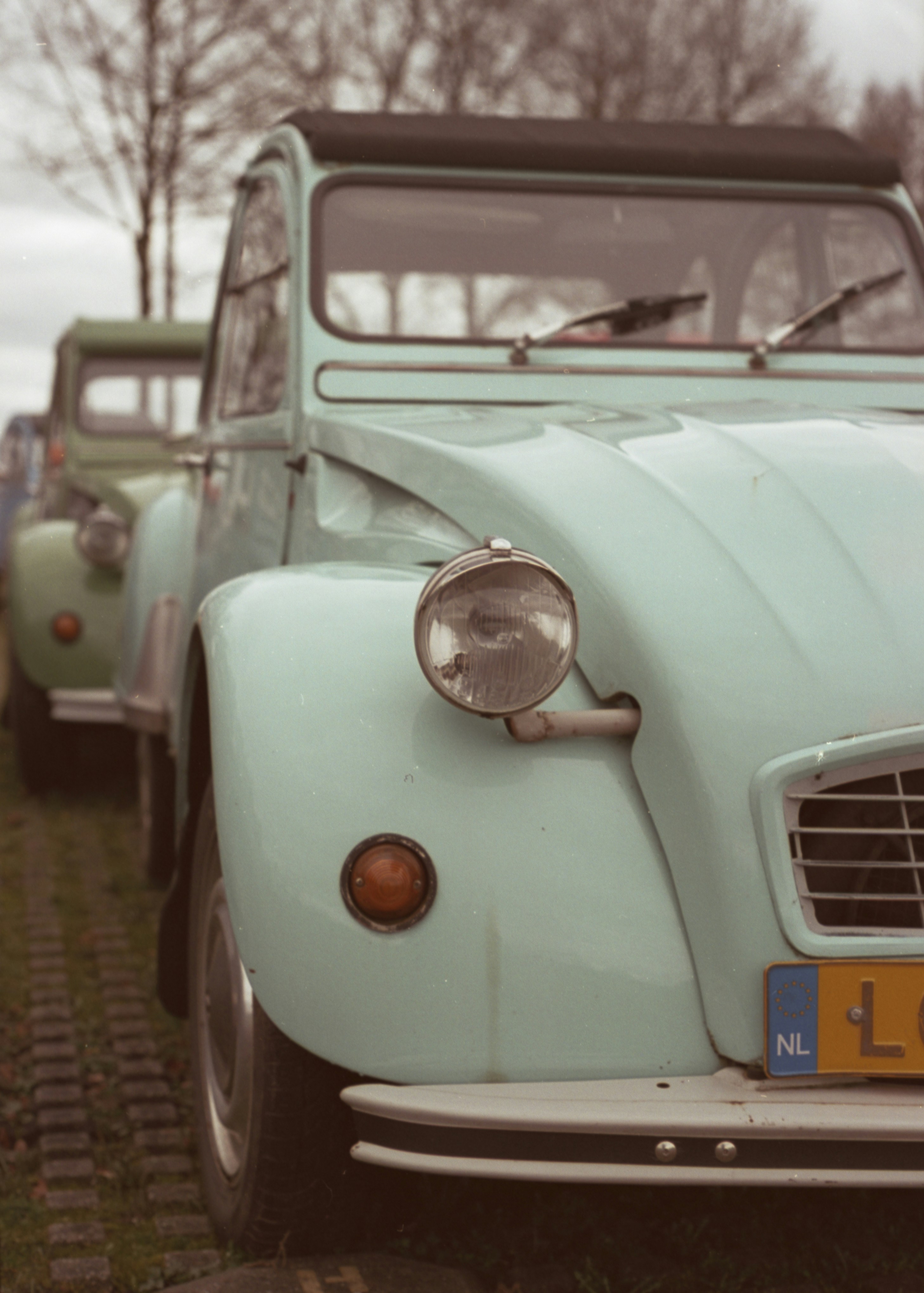 Mint turquoise vintage car with a prominent front headlight, parked among a line of similar vehicles; a nostalgic auto show scene with soft blur and overcast sky.
