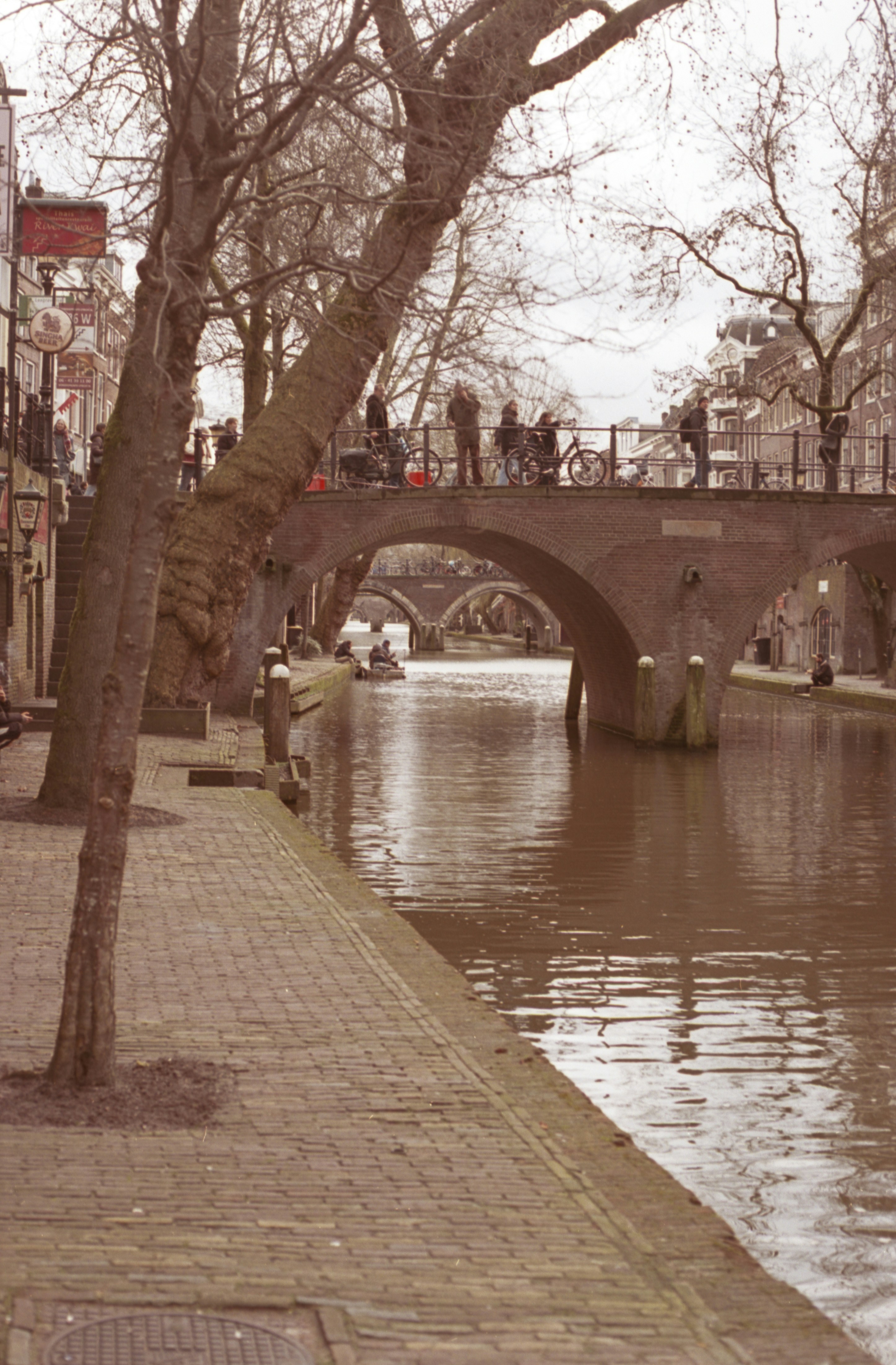 Muted canal scene with a brick bridge and pedestrians, bicycles, and boats along the waterway. A tree-lined path runs beside the canal, giving a quiet, urban-ramble feel. (Note: this is a photographic capture, not a drawing or illustration).