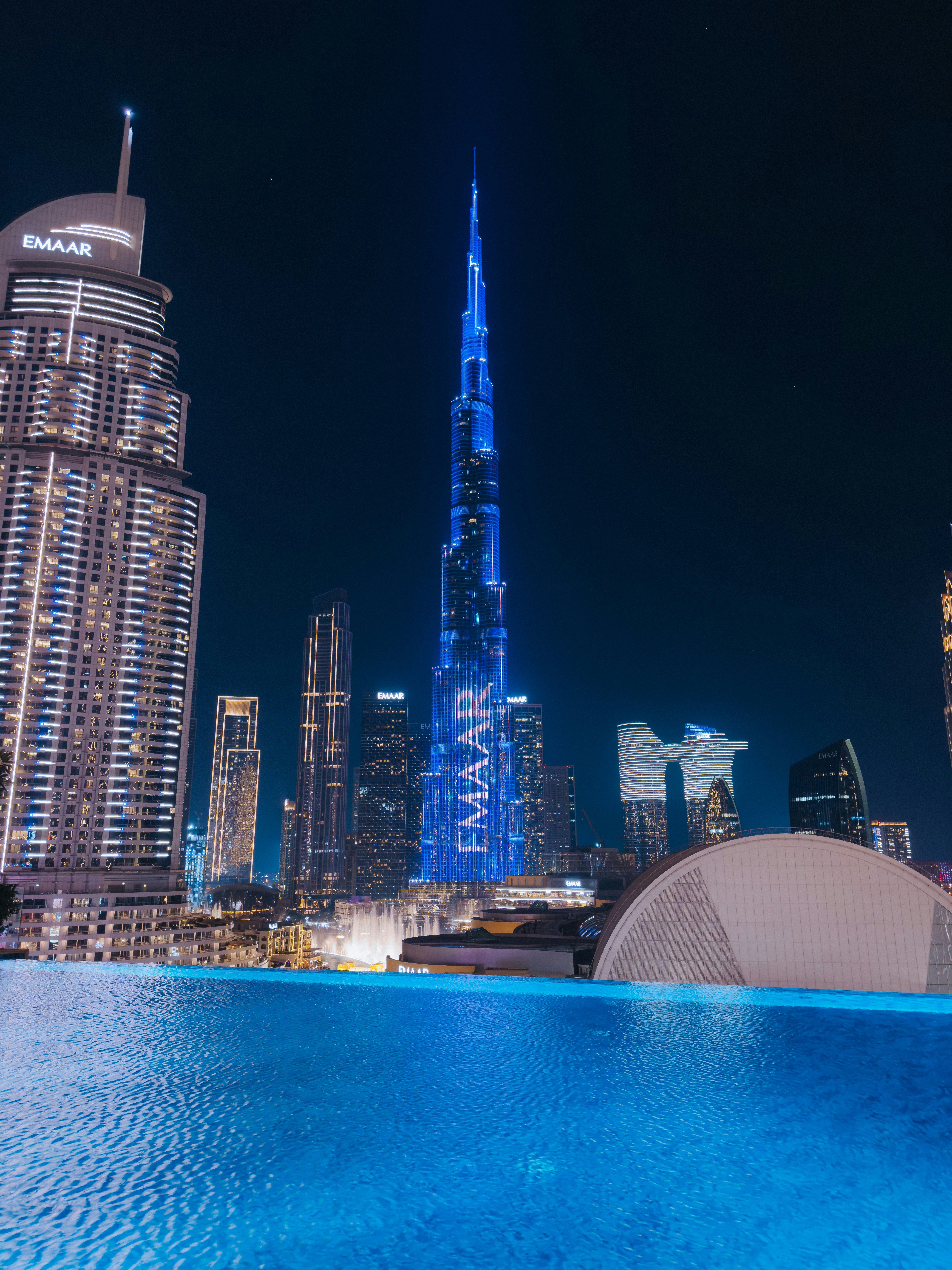 Blue-lit Burj Khalifa and surrounding towers rise above a glowing waterfront, with the pool foreground reflecting city lights.
