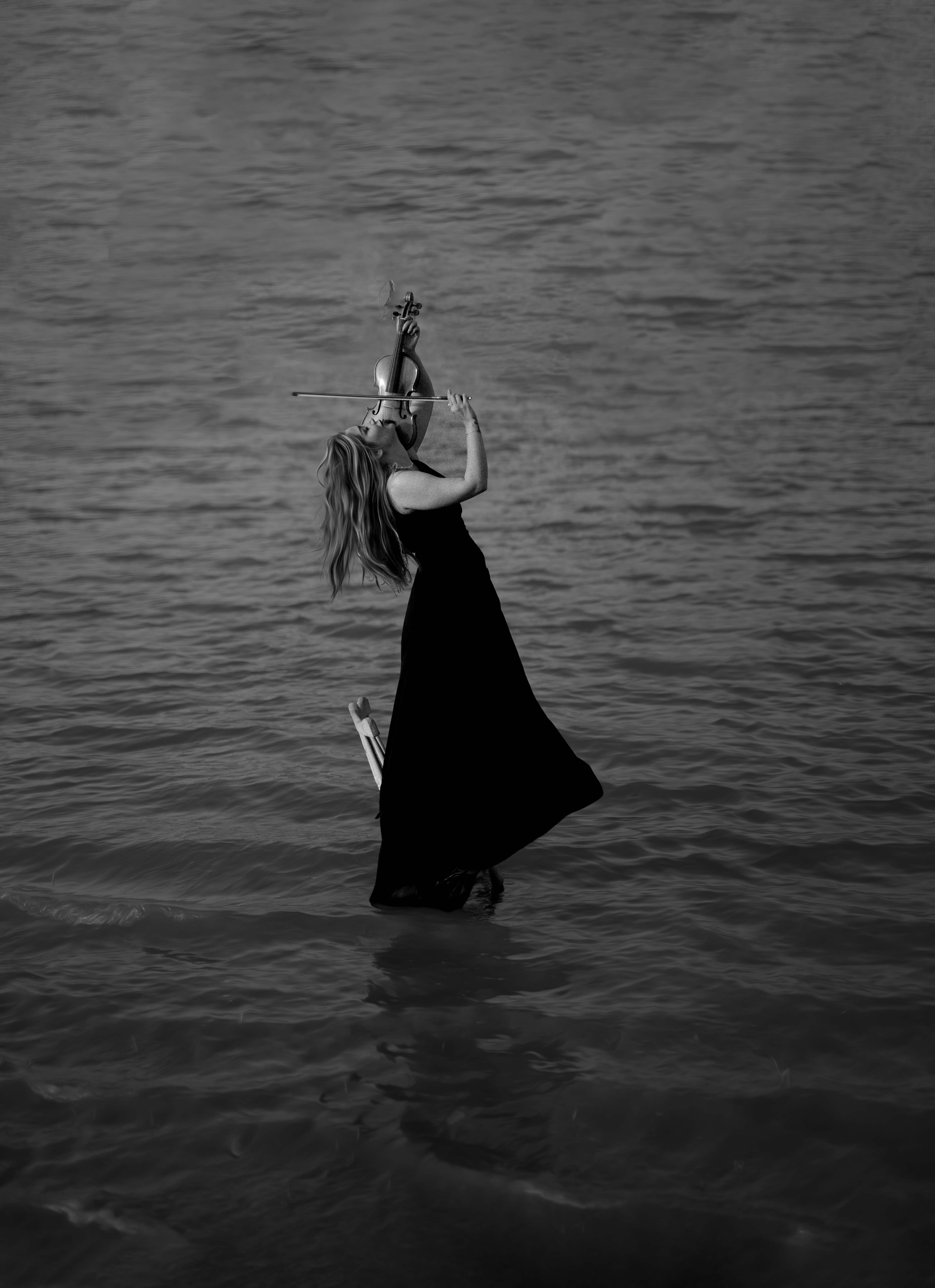 A lone woman in a flowing black gown stands knee-deep in dark water, balancing a long rod across her hands as waves ripple around her. The scene is stark and moody, emphasizing posture and form in monochrome.
