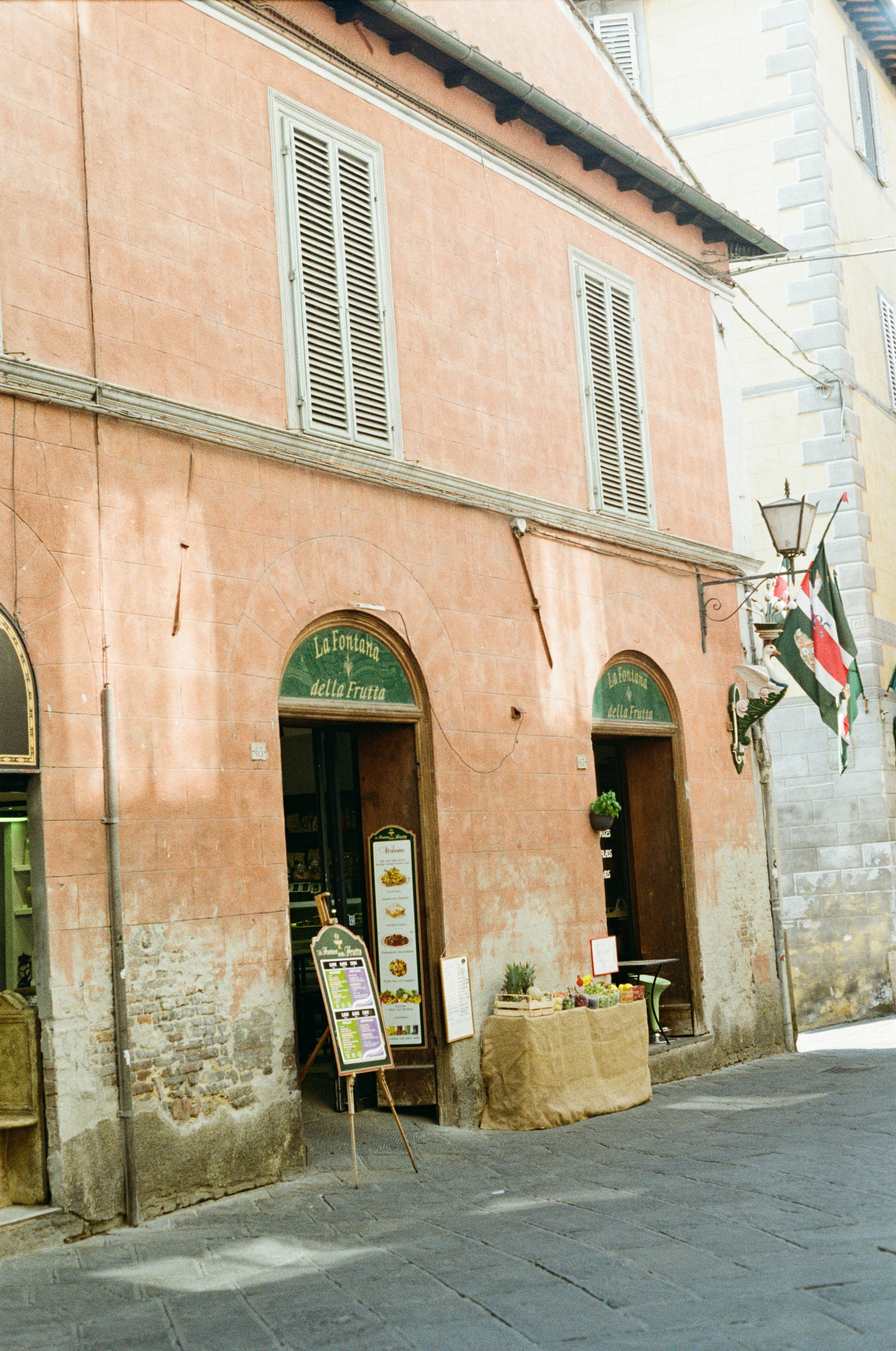 A terracotta-pink building houses a greengrocer with menu boards and a flag over the quiet street.