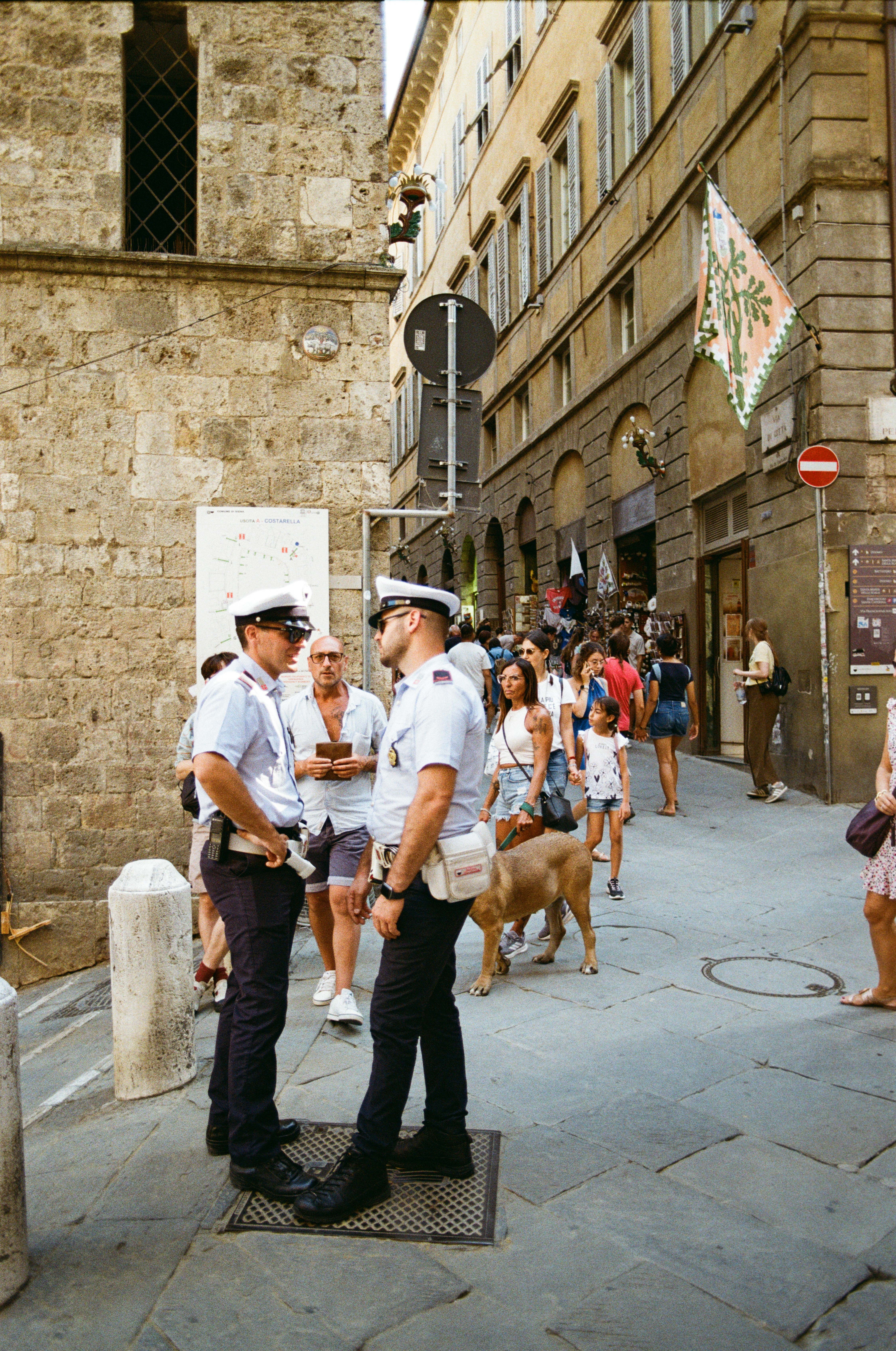 Bustling street scene with two uniformed officers conversing near a stone arch, pedestrians and a dog in the background; a lively urban moment in a historic Italian square.