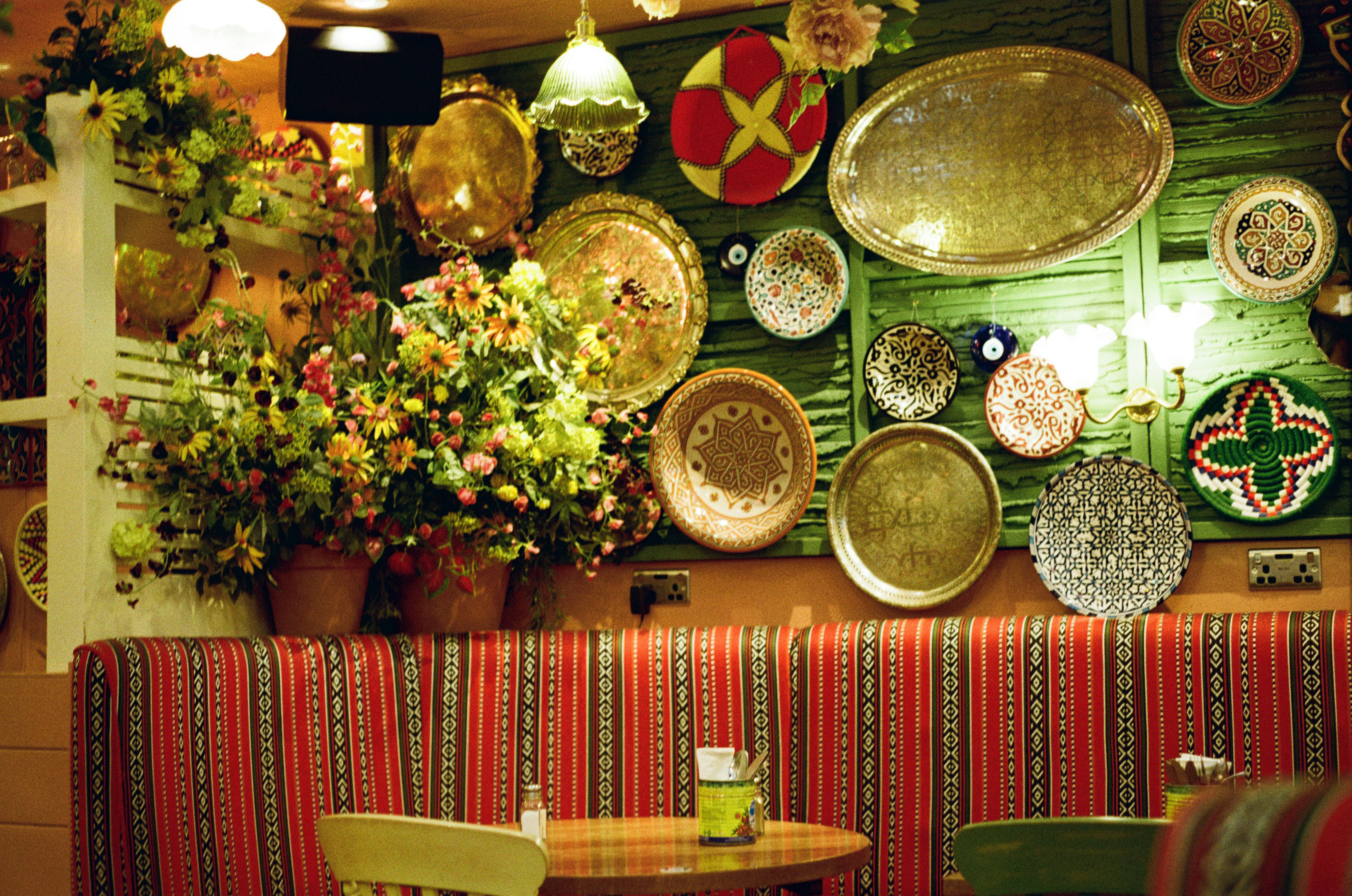 Colorful plates adorn a green wall above a striped booth with a bouquet of flowers and a small round table, creating a warm, nostalgic dining scene.