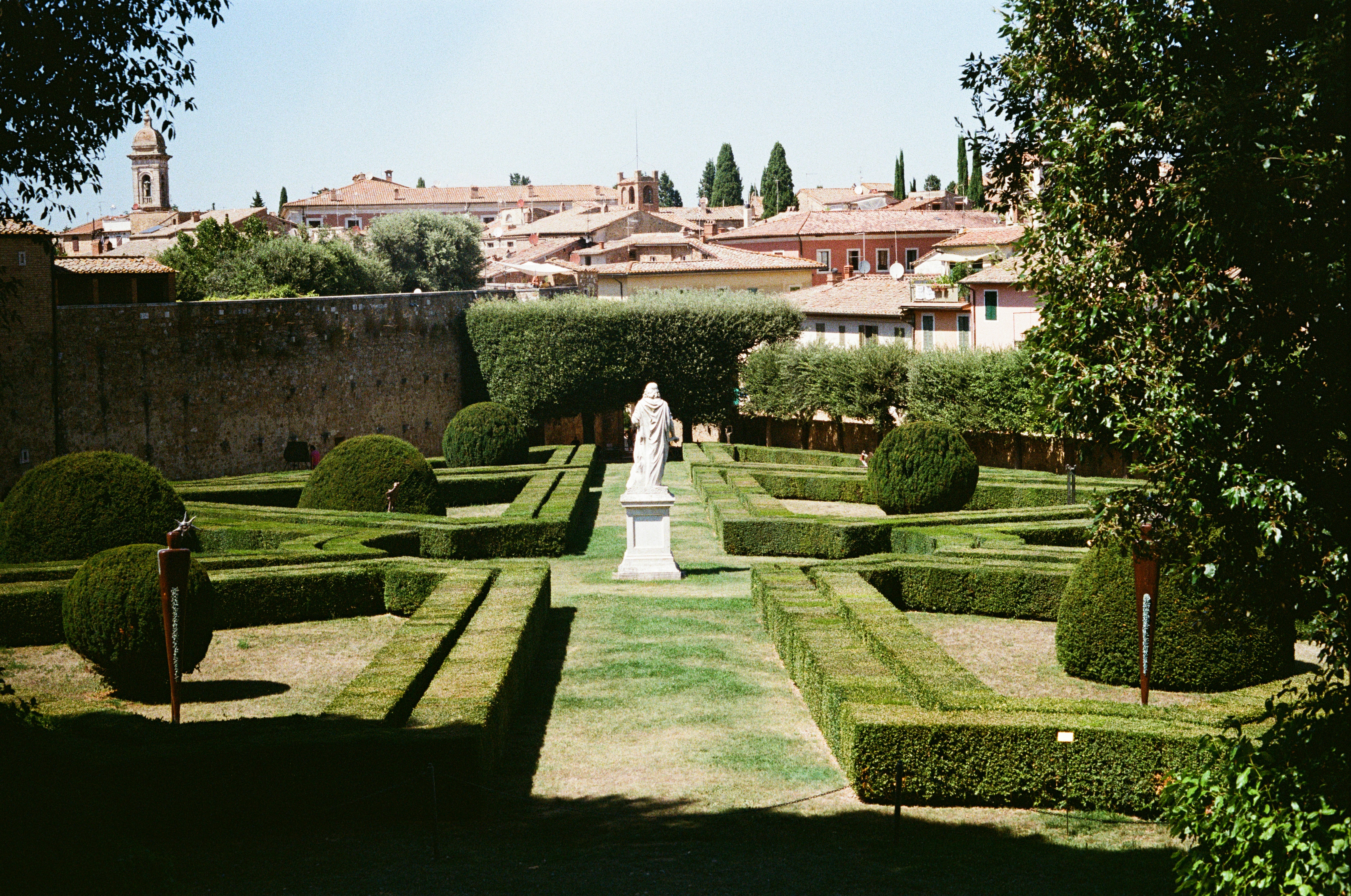 Manicured classical garden with a central statue framed by hedges and symmetry, revealing a distant townscape beyond the garden walls.