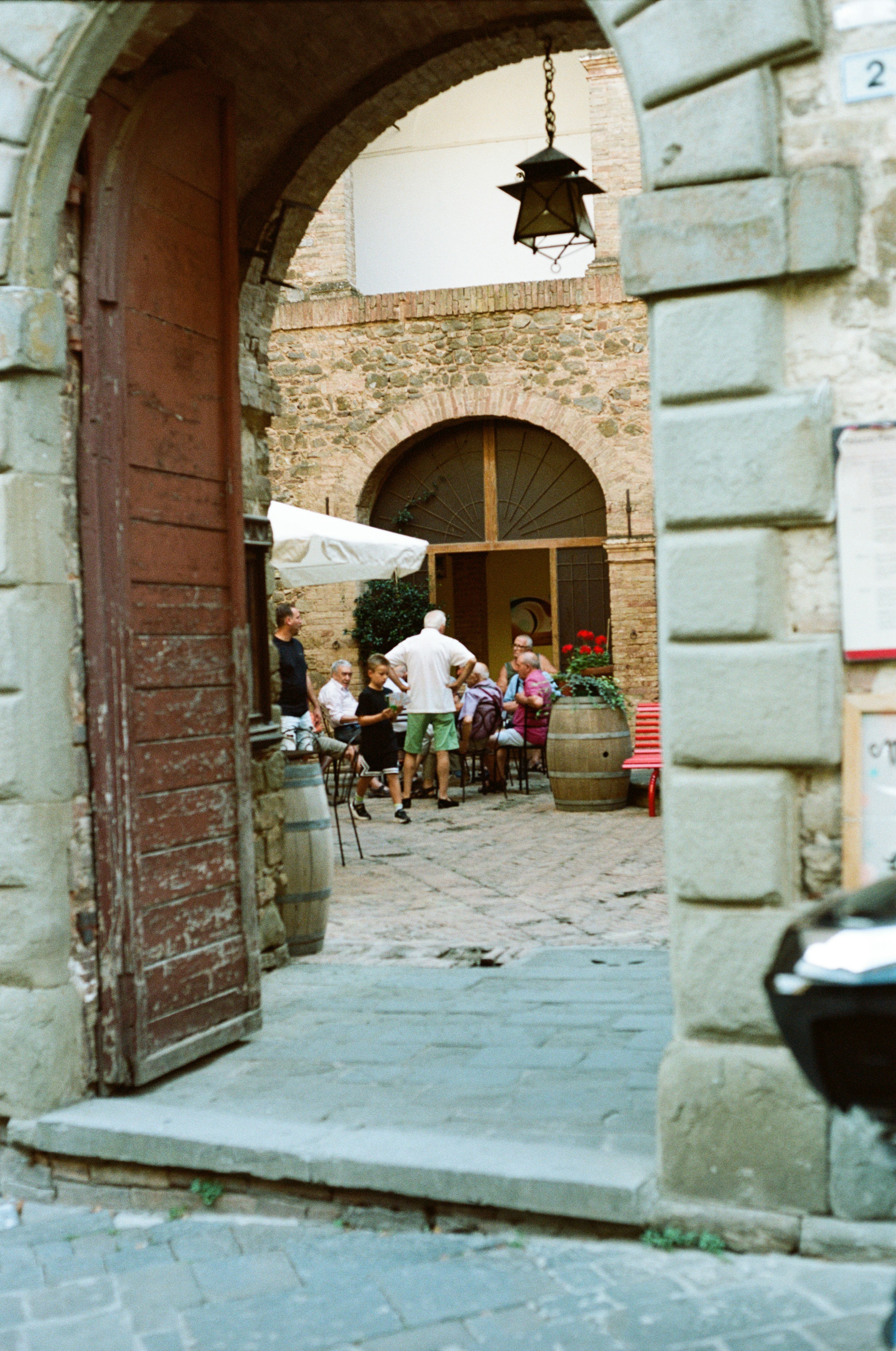 Bustling courtyard scene framed by a stone arch, with locals and visitors gathered near a doorway under an umbrella. A rustic, sunny setting with outdoor seating and warm brick tones.