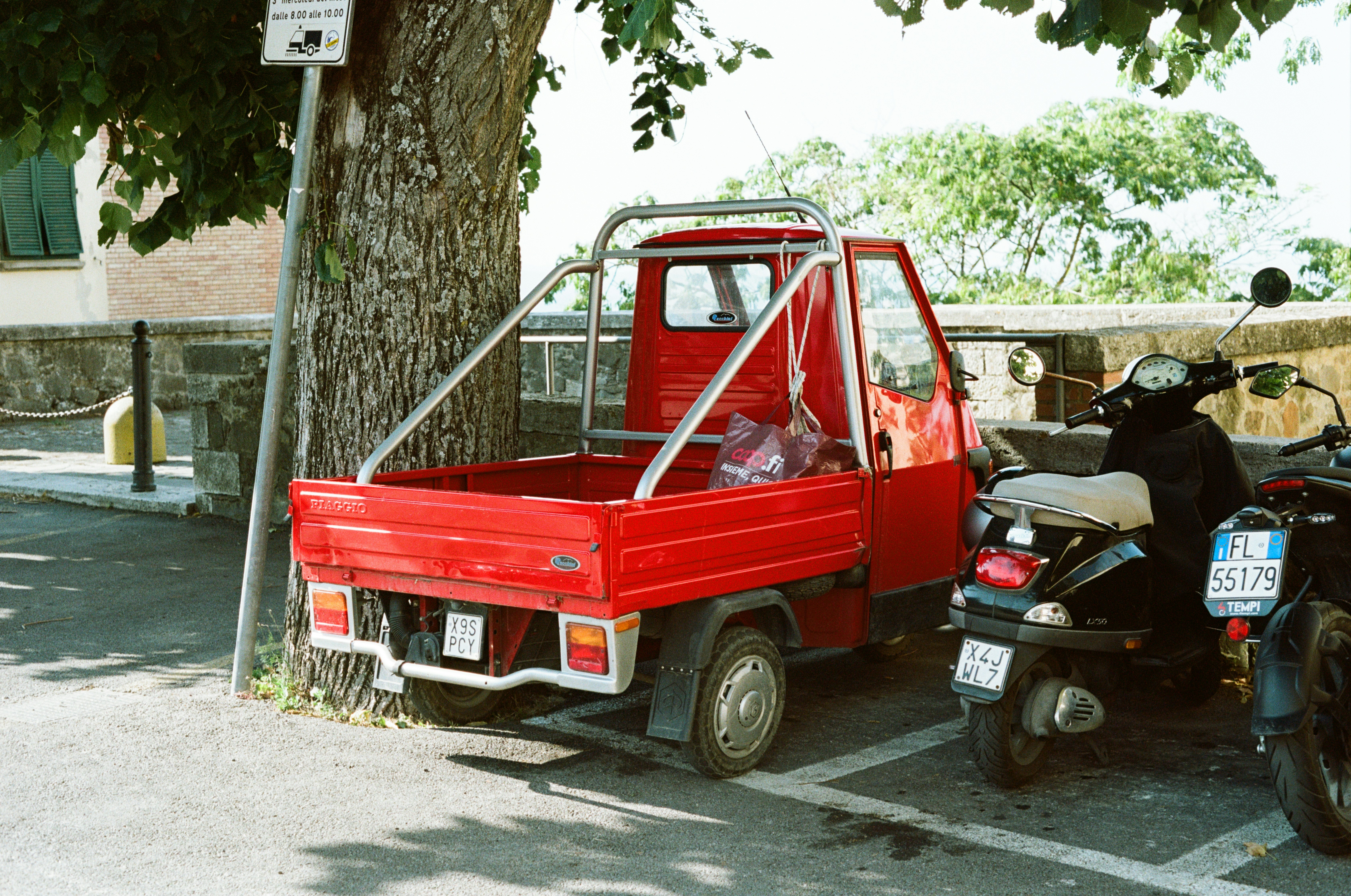 Red utility truck parked beside a scooter under a tree, in a sunlit street scene with a parked sign and greenery in the background.