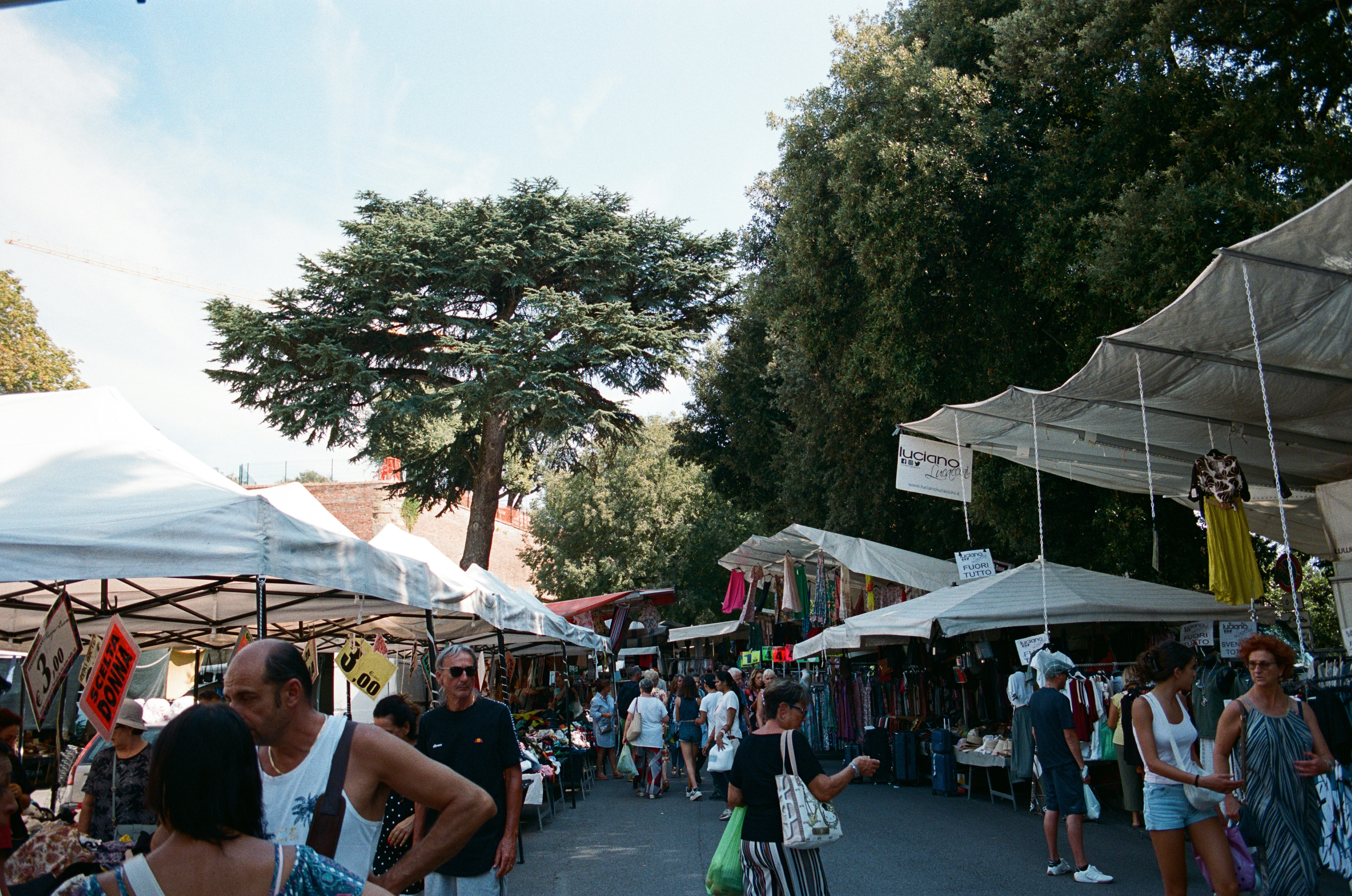 Bustling outdoor market with white tents, shoppers, and vendors along a sunlit street.