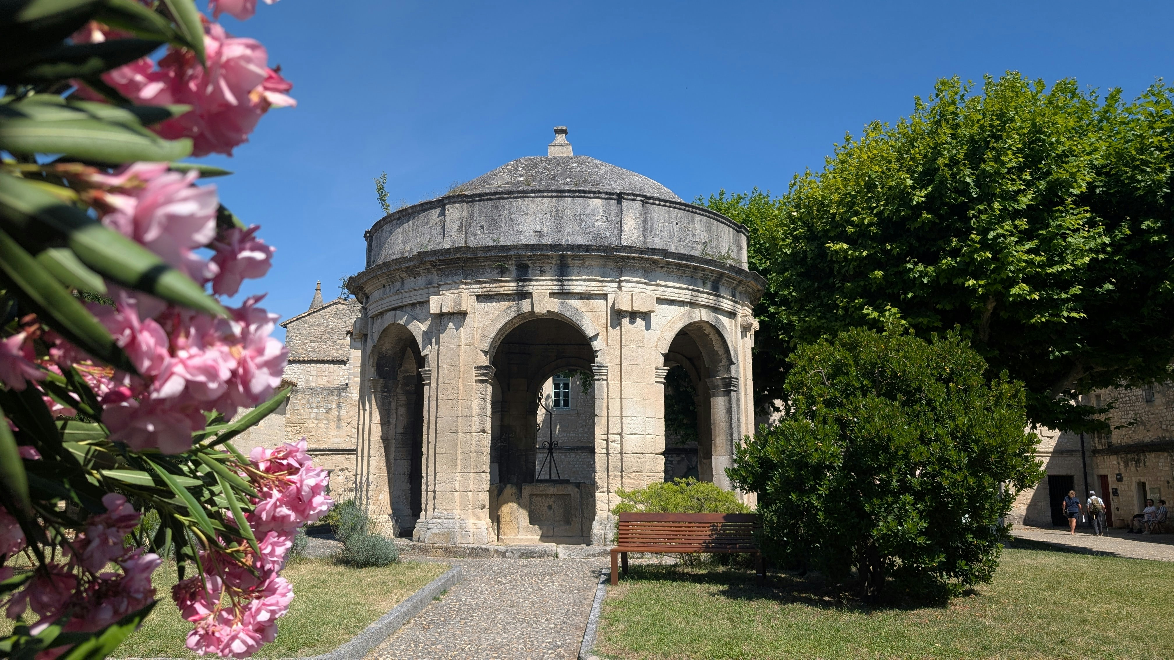 A circular stone pavilion with arches sits at the end of a gravel path, framed by lush greenery and pink blossoms in the foreground.
