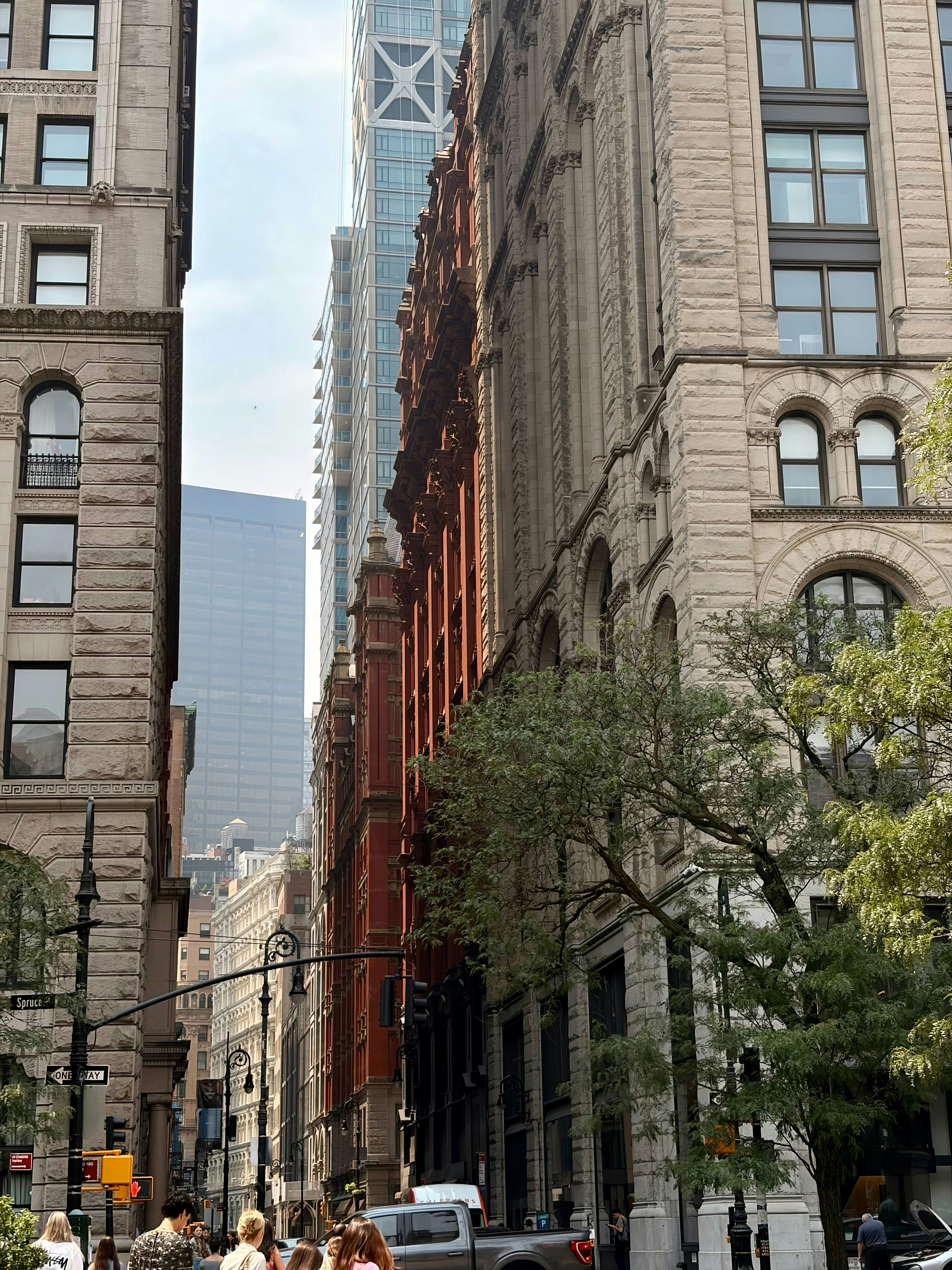 A street-level urban scene with tall historic buildings lining a narrow avenue, modern glass towers peeking above, and pedestrians at the curb. A mix of architectural textures and greenery frames the busy city corridor.