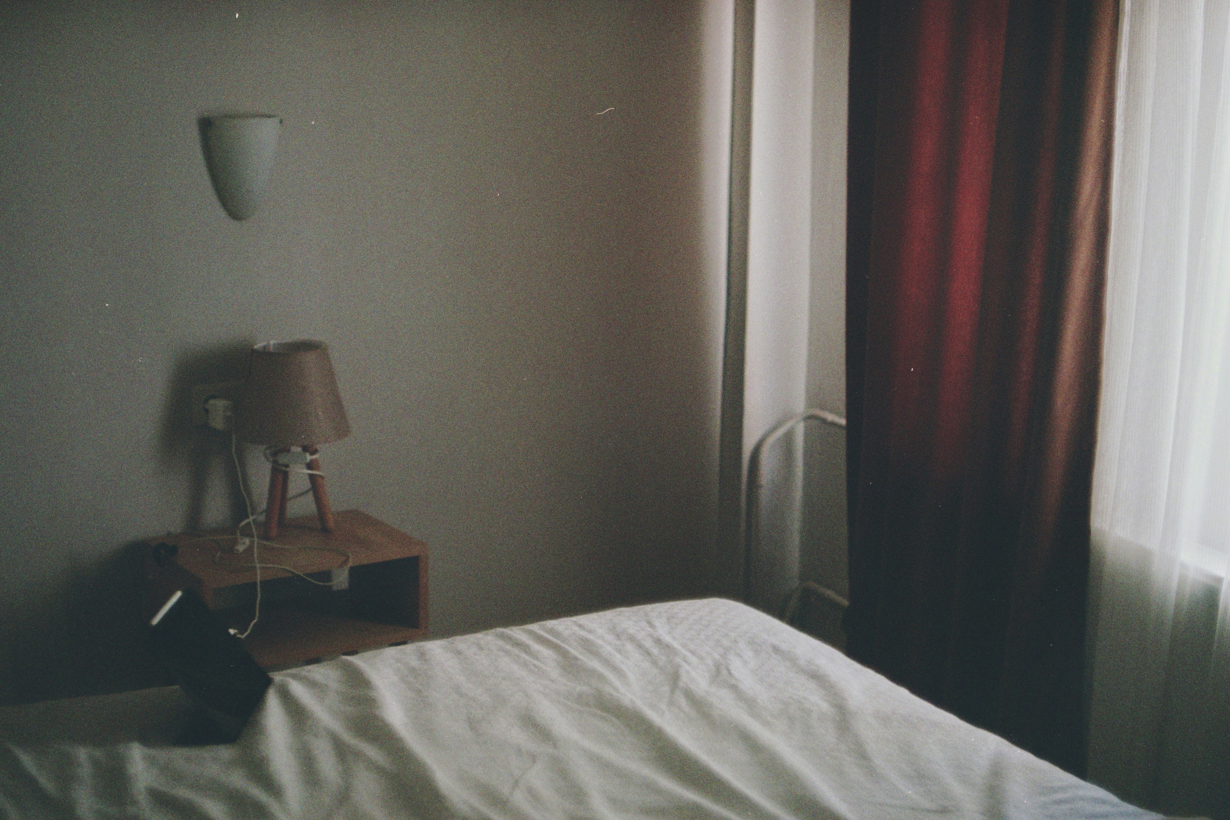 Dimly lit hotel bedroom with a neatly made bed, a nightstand, and a small lamp; curtains partially drawn, soft natural light mid-morning. A simple, moody interior scene.