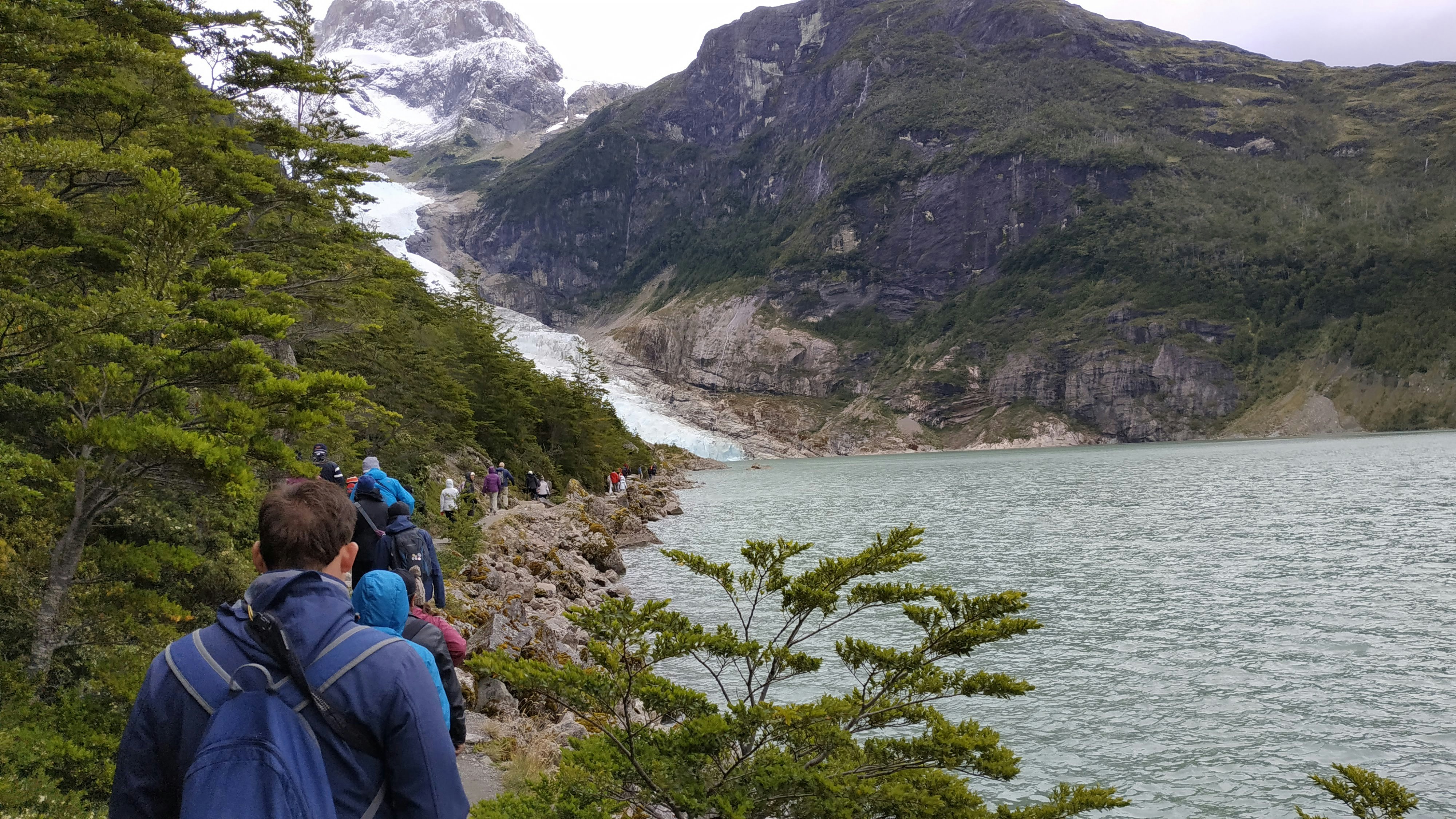 Trail of hikers follows a rocky shoreline beside a glacial lake with distant mountains; a forest edge frames the foreground, suggesting a remote alpine hike.