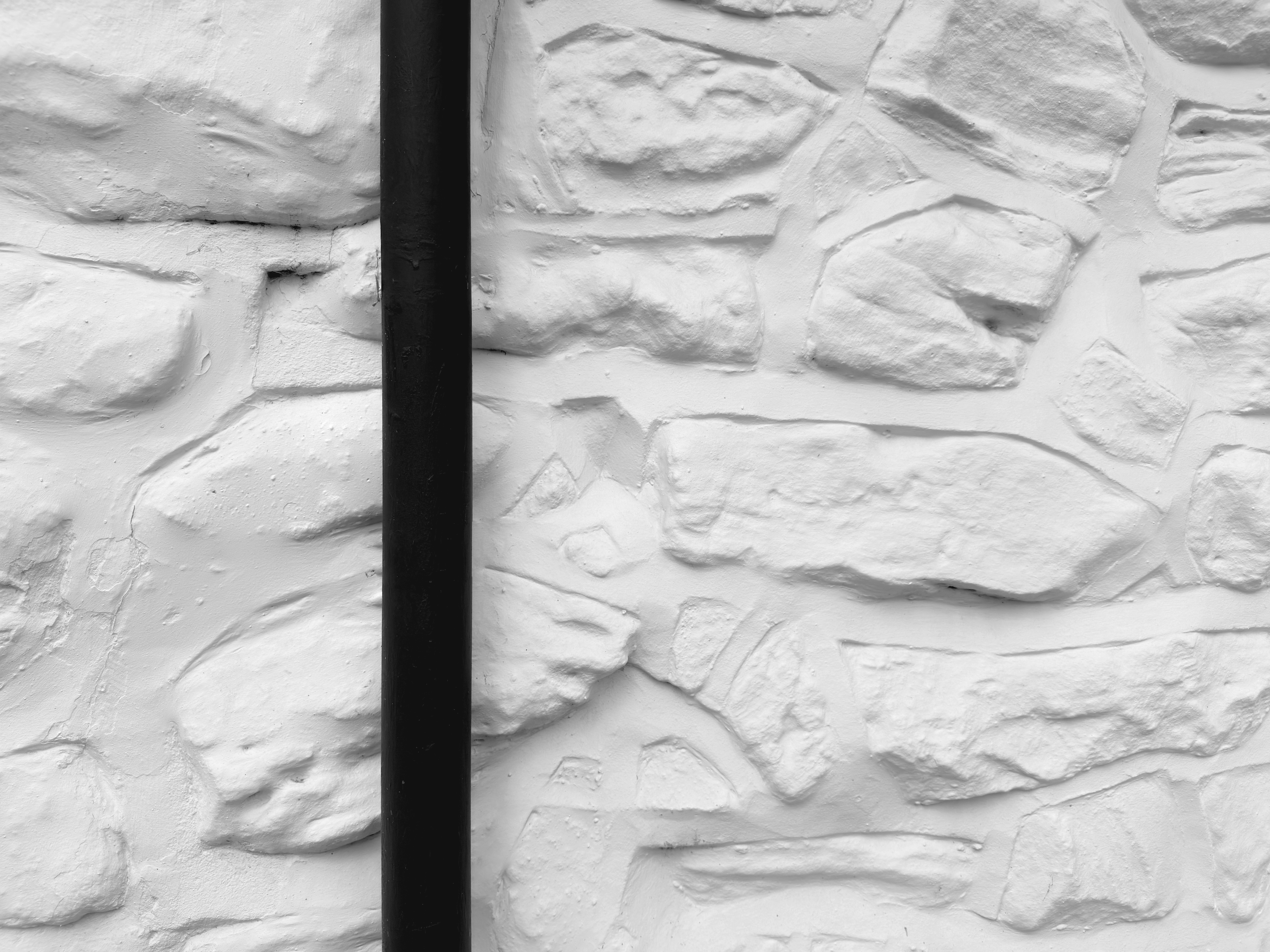 Monochrome stones forming a textured wall with a dark vertical pipe on the left side, creating strong architectural contrast.
