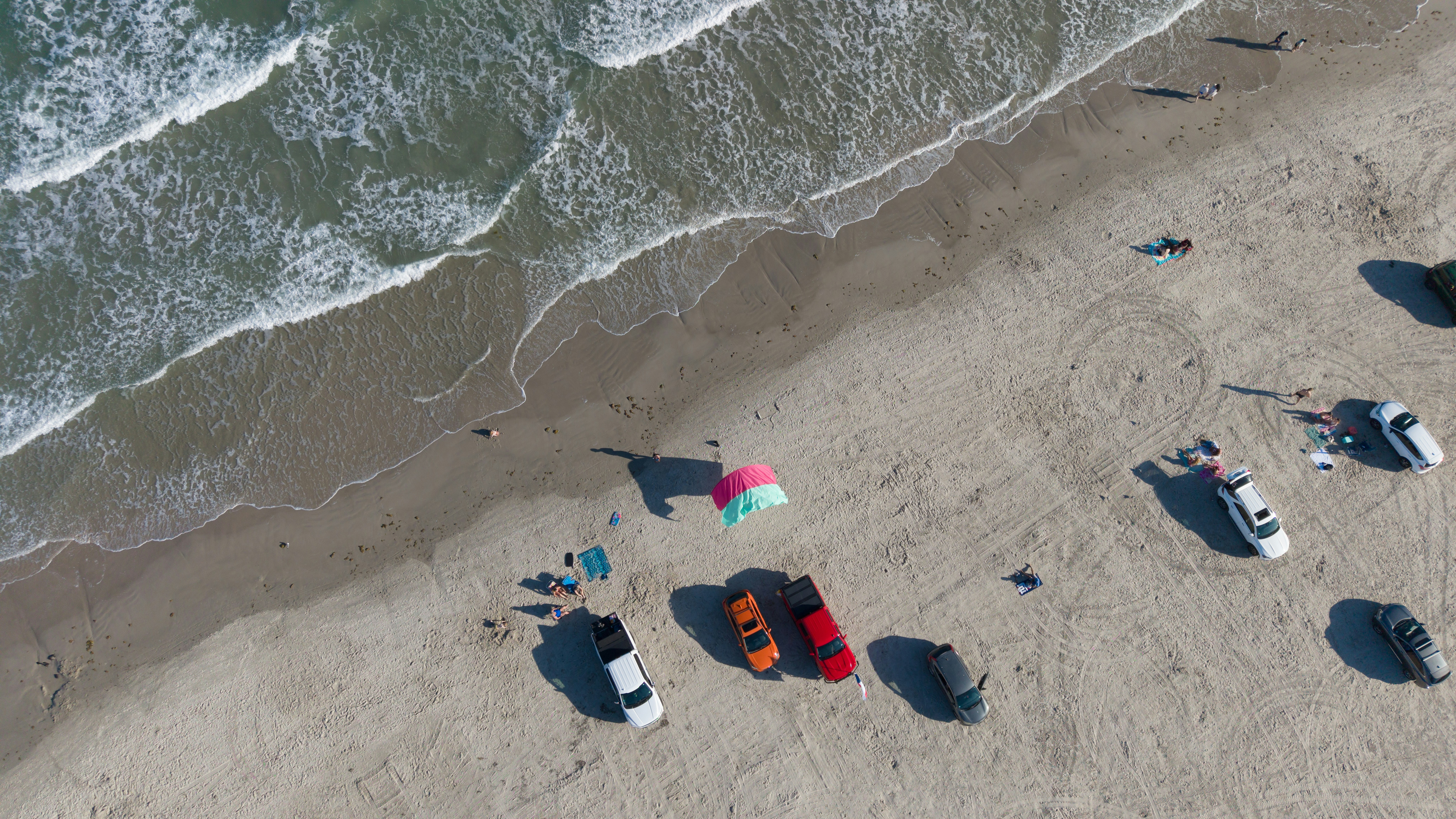 Aerial view of a sandy beach with parked vehicles near the waterline, a colorful umbrella casting a shadow, and small groups of people along the shore.