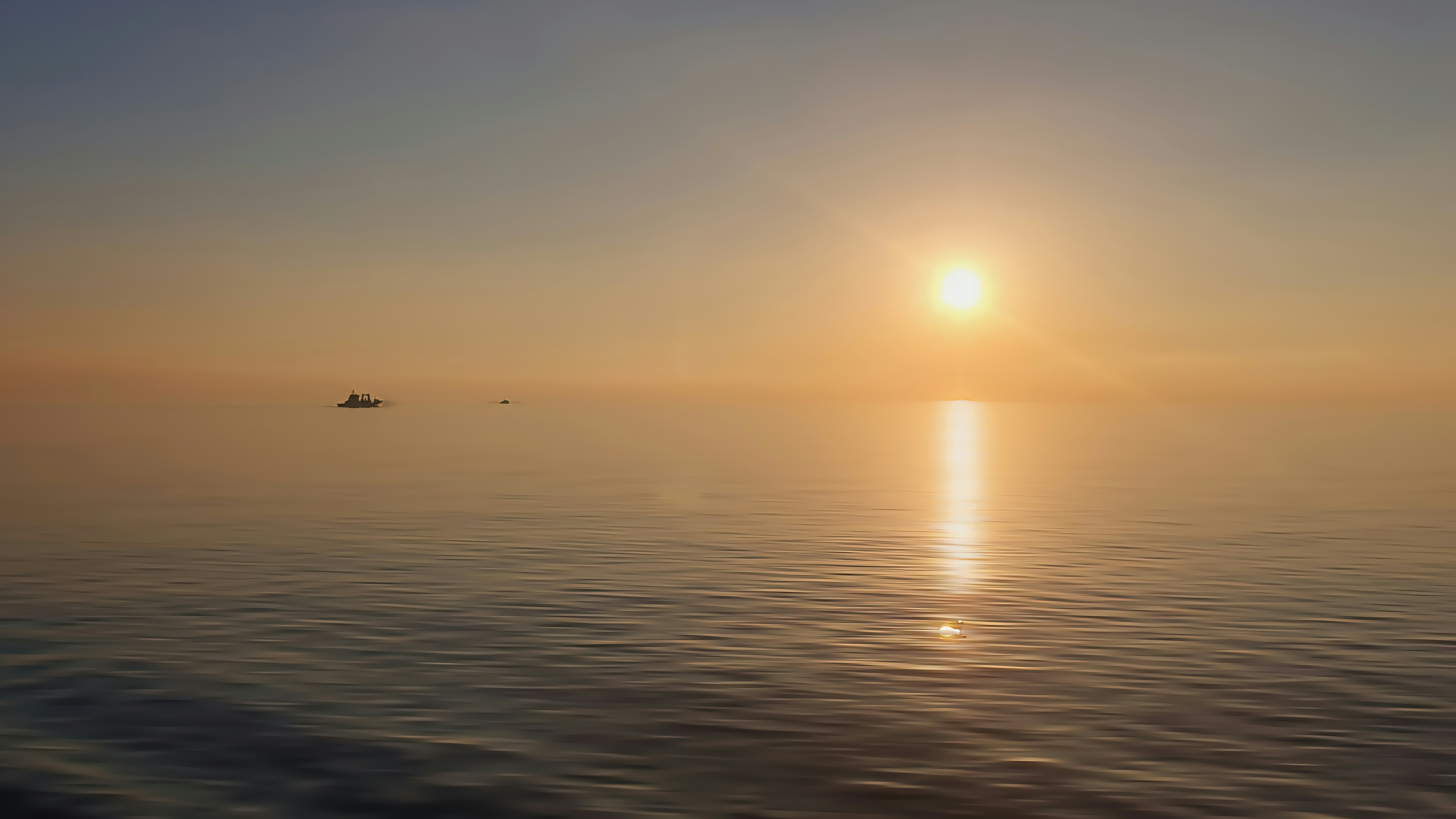 Sunlit horizon over a calm sea with a small distant vessel on the left; warm gradient sky and gentle waves.