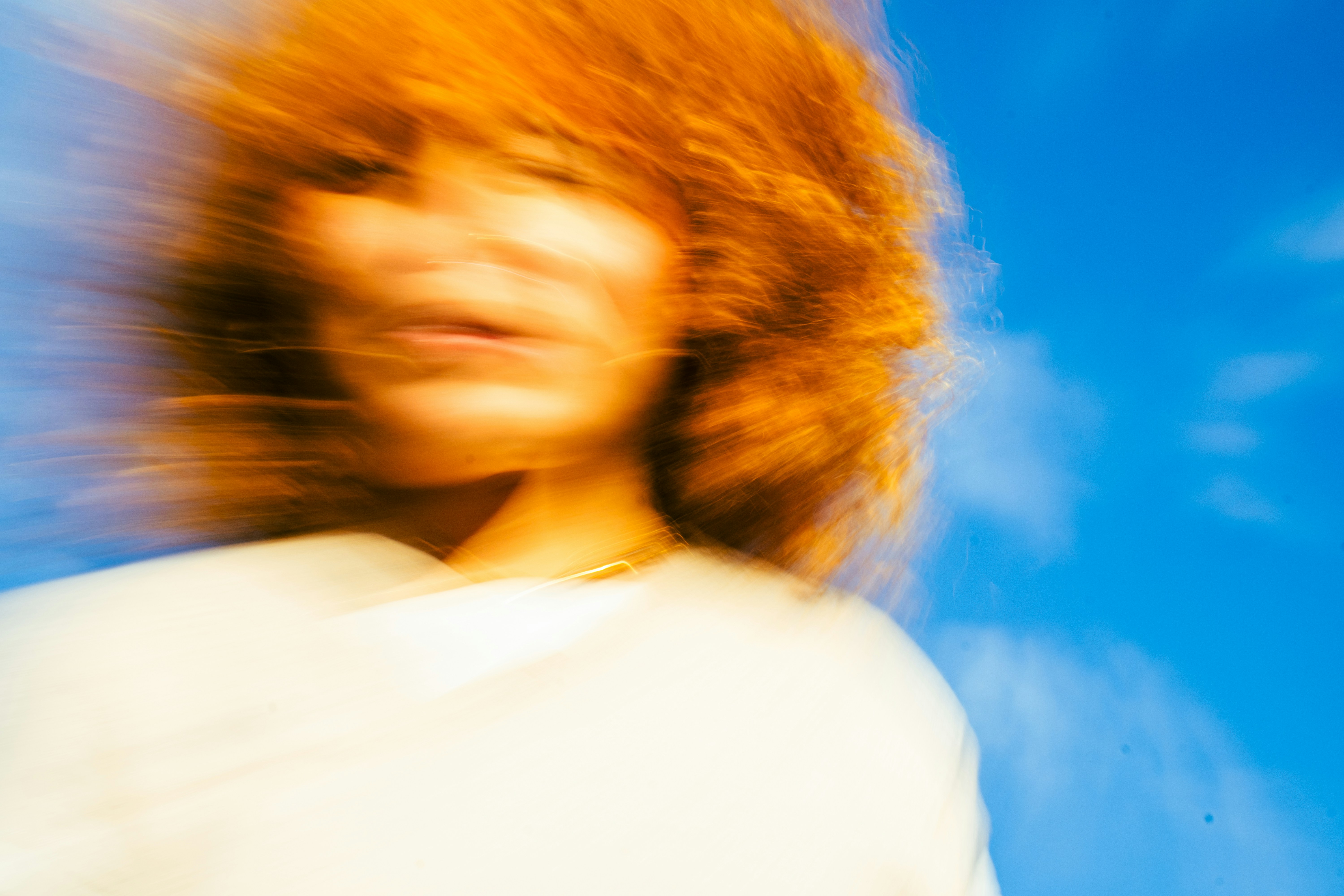 A motion-blurred portrait of a person with warm hair against a bright blue sky, emphasizing color and motion over facial detail. This is a photographic capture rather than a painting or illustration.