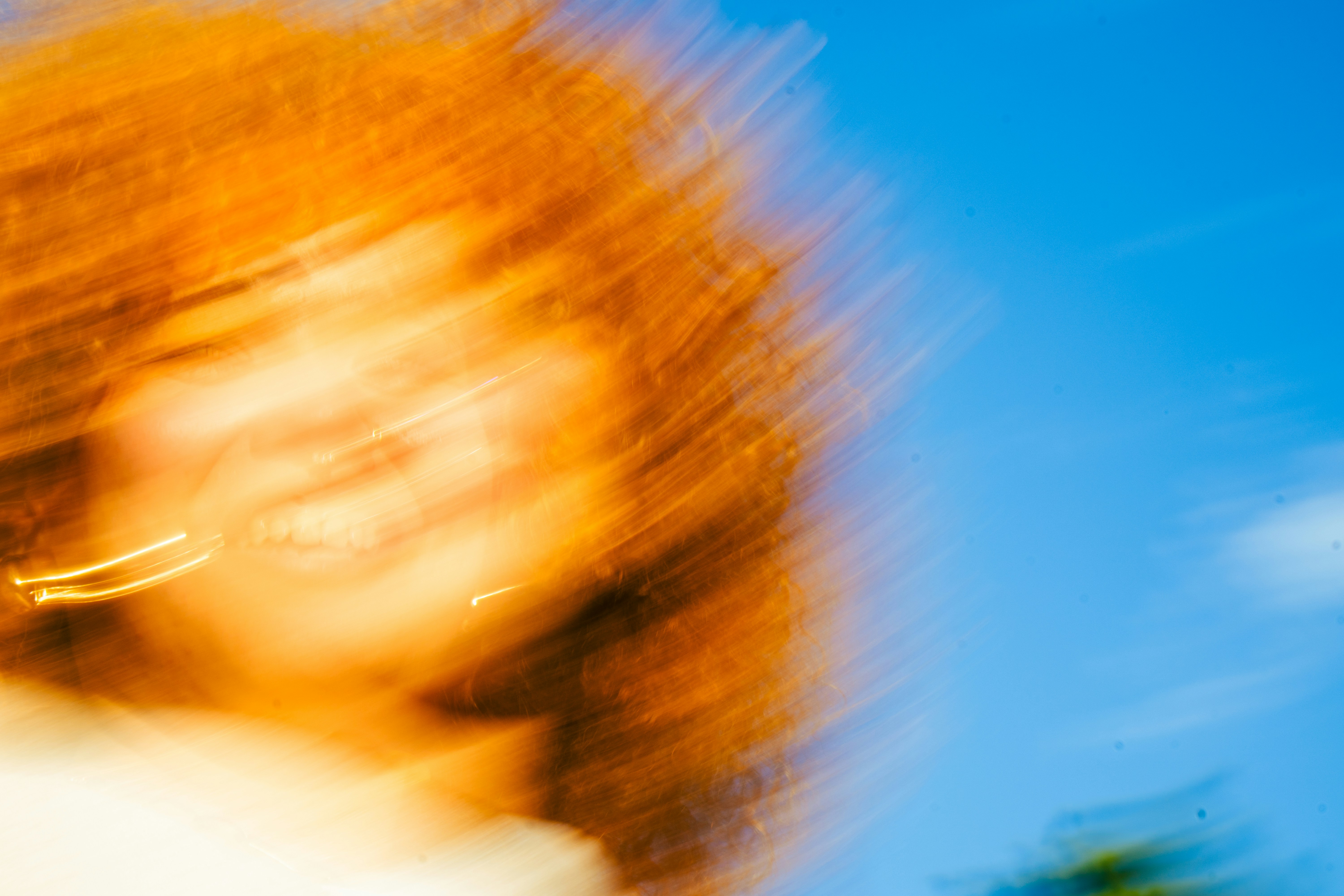 Sunlit blur of a person with curly hair against a bright blue sky; motion creates abstract warmth and energy. A candid, dynamic snapshot with strong color contrast. This is a real photograph, not a drawing or painting.
