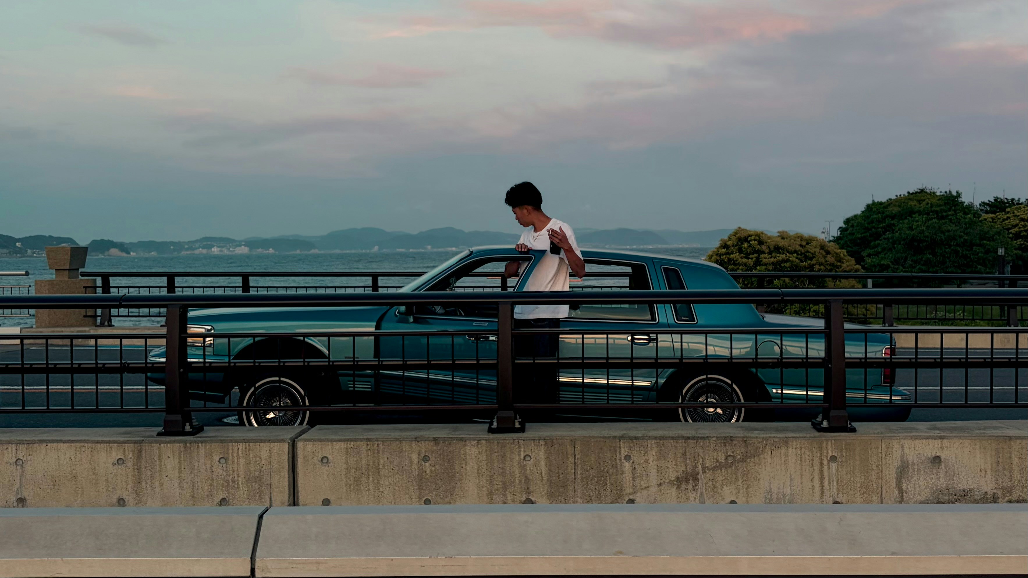A man stands next to a teal vintage car along a waterfront railing; shallow depth emphasizes him as the focal figure against a calm bay and distant hills.
