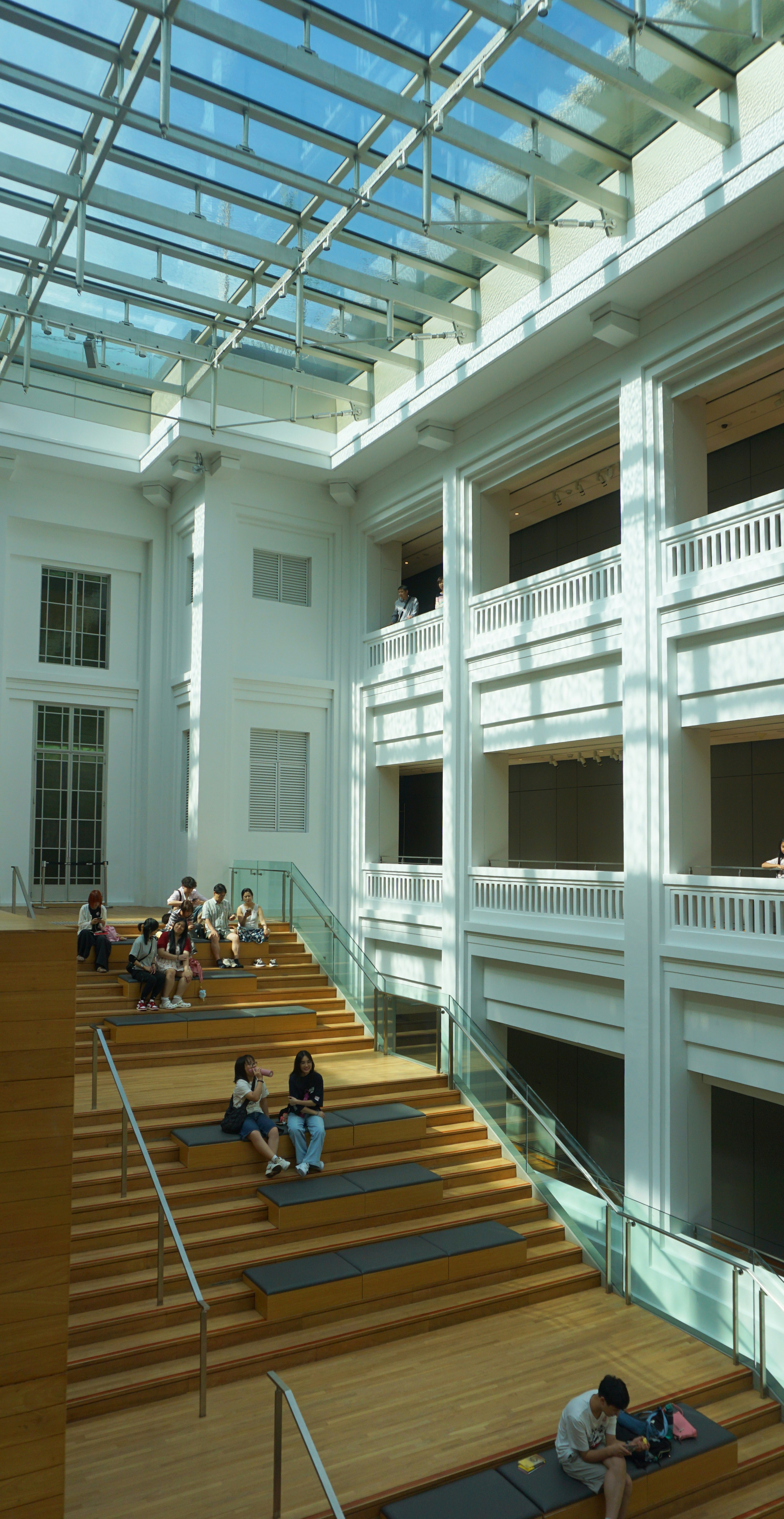 Sunlit Concourse: People Gather on Wide Wooden Steps Inside a Glass-Canopied Atrium