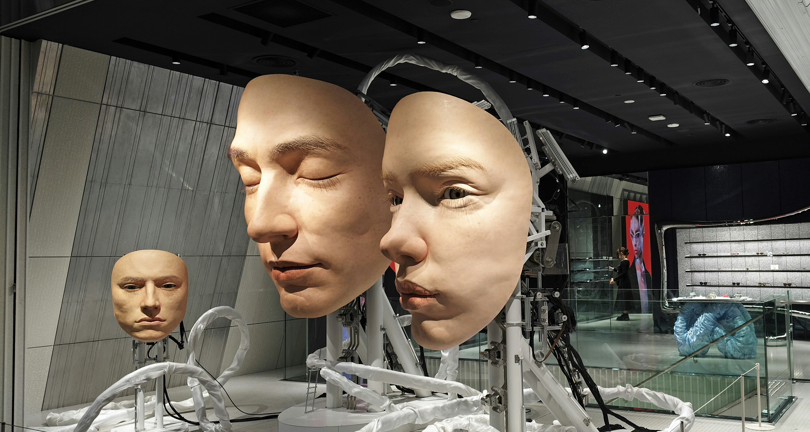 Three large sculpted faces suspended in a futuristic gallery space, with metallic support structures and a red-screen exhibit in the background.