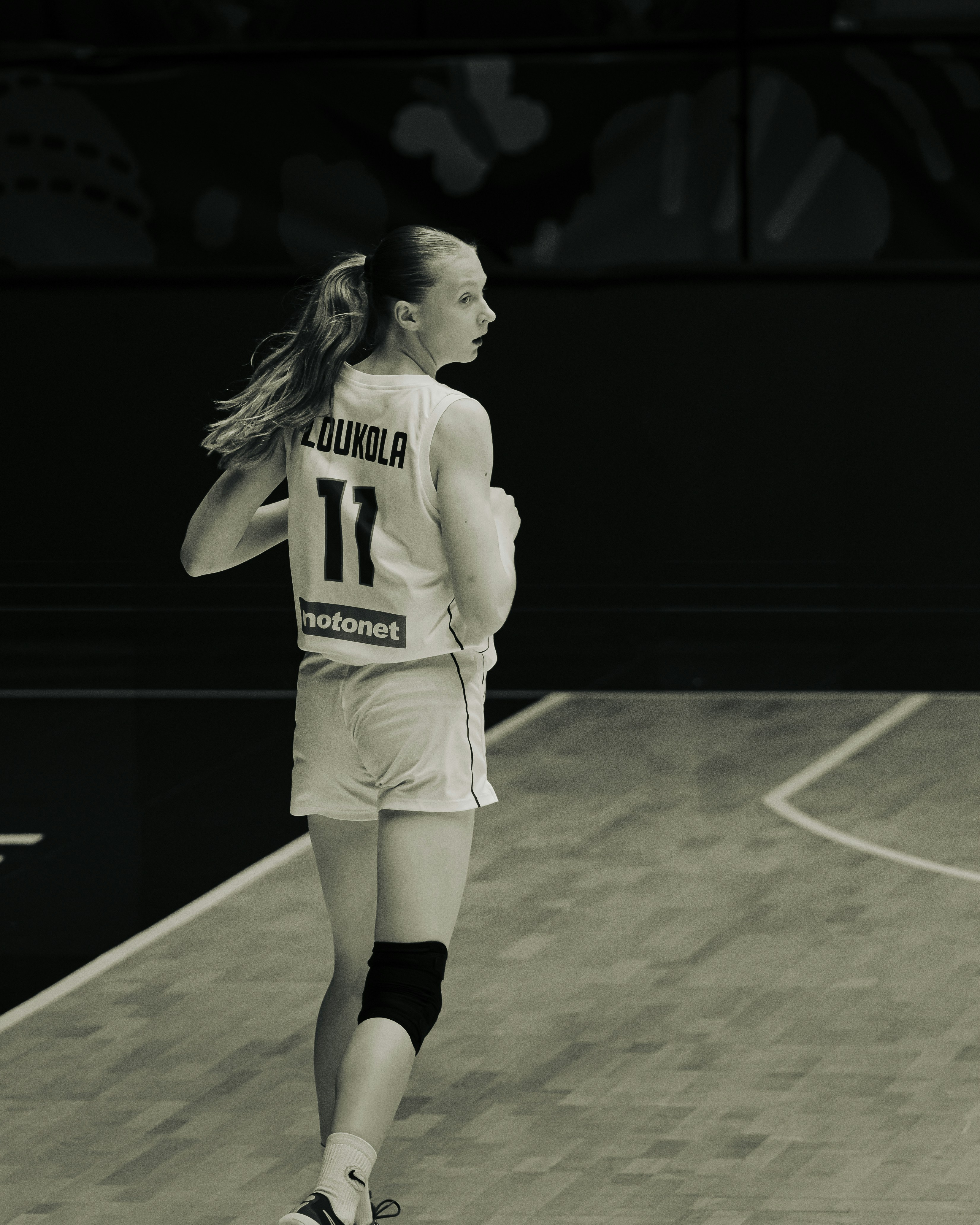 Monochrome action of a female basketball player in uniform with number 11, turning on the court; leg knee wrap and sponsor patch visible, foreground subject framed against dark background.