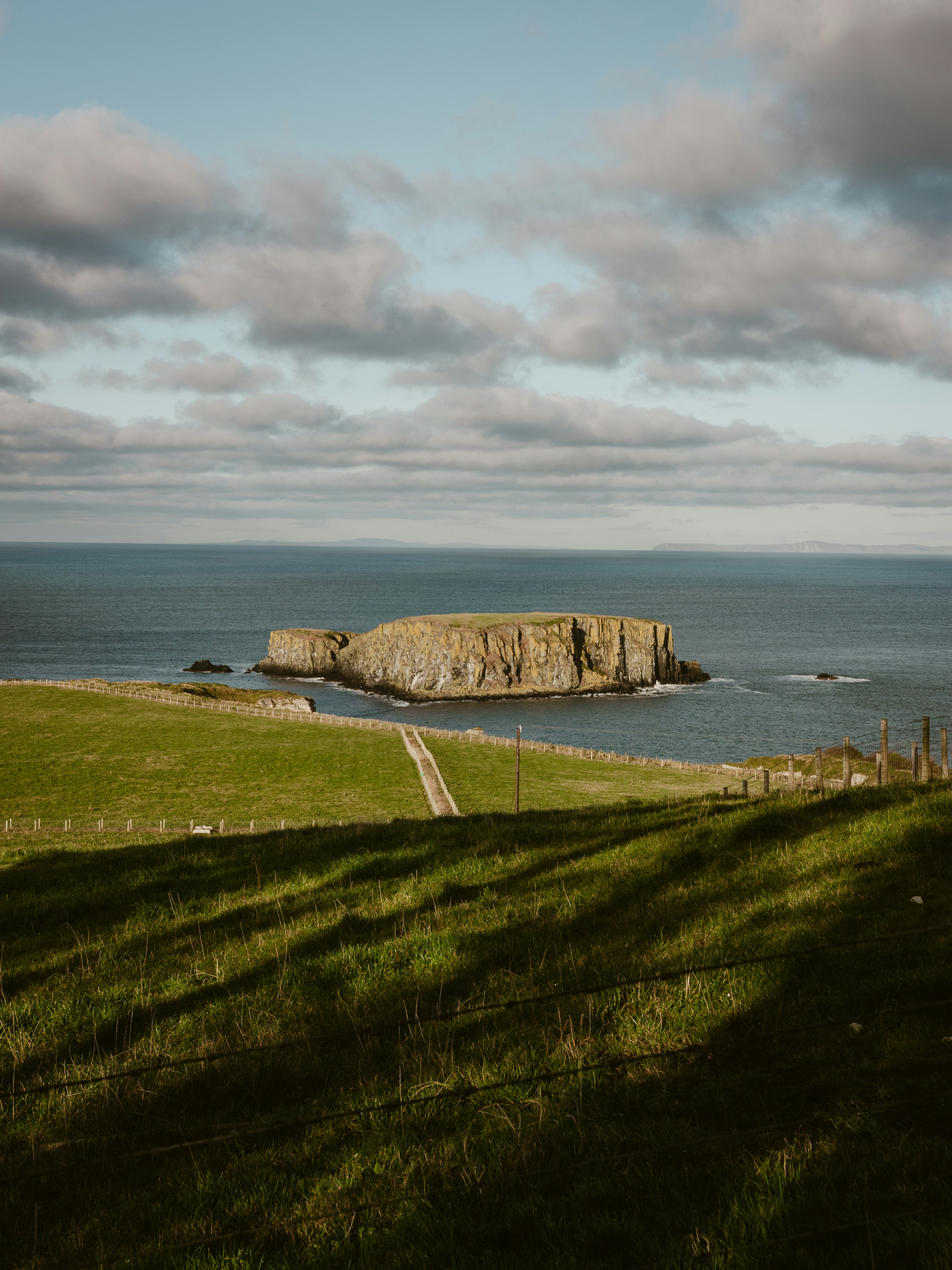 Stone sea stack rising from calm blue water with grassy cliffs in the foreground and a fence line under a partly cloudy sky. A tranquil coastal scene emphasizing open space and natural textures.